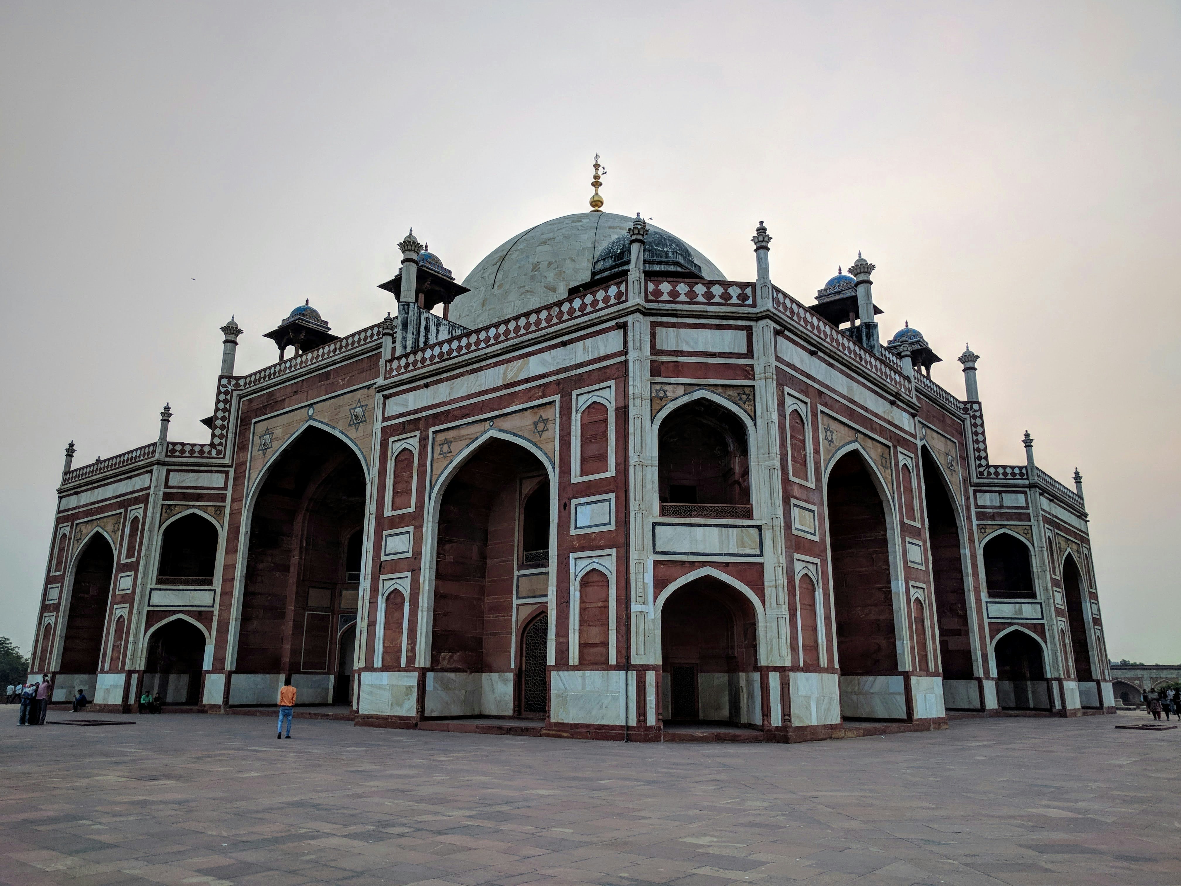 Red sandstone Mughal-era facade with a central blue-gray dome and repeating arched gateways, set in a spacious plaza.
