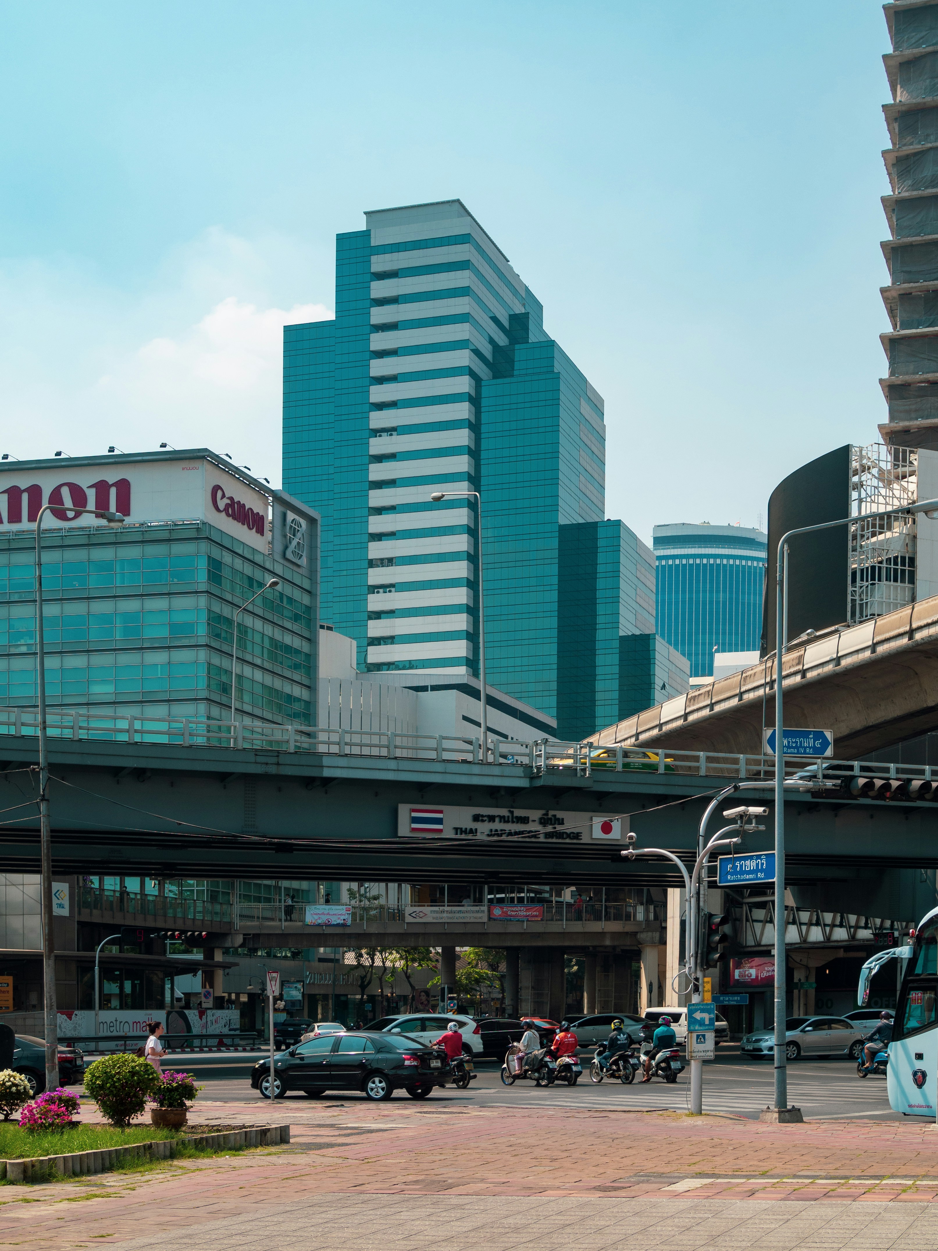 A vibrant cityscape showcasing modern architecture with a Canon logo prominently displayed, capturing the essence of urban life. Traffic flows beneath elevated train tracks amidst towering skyscrapers.
