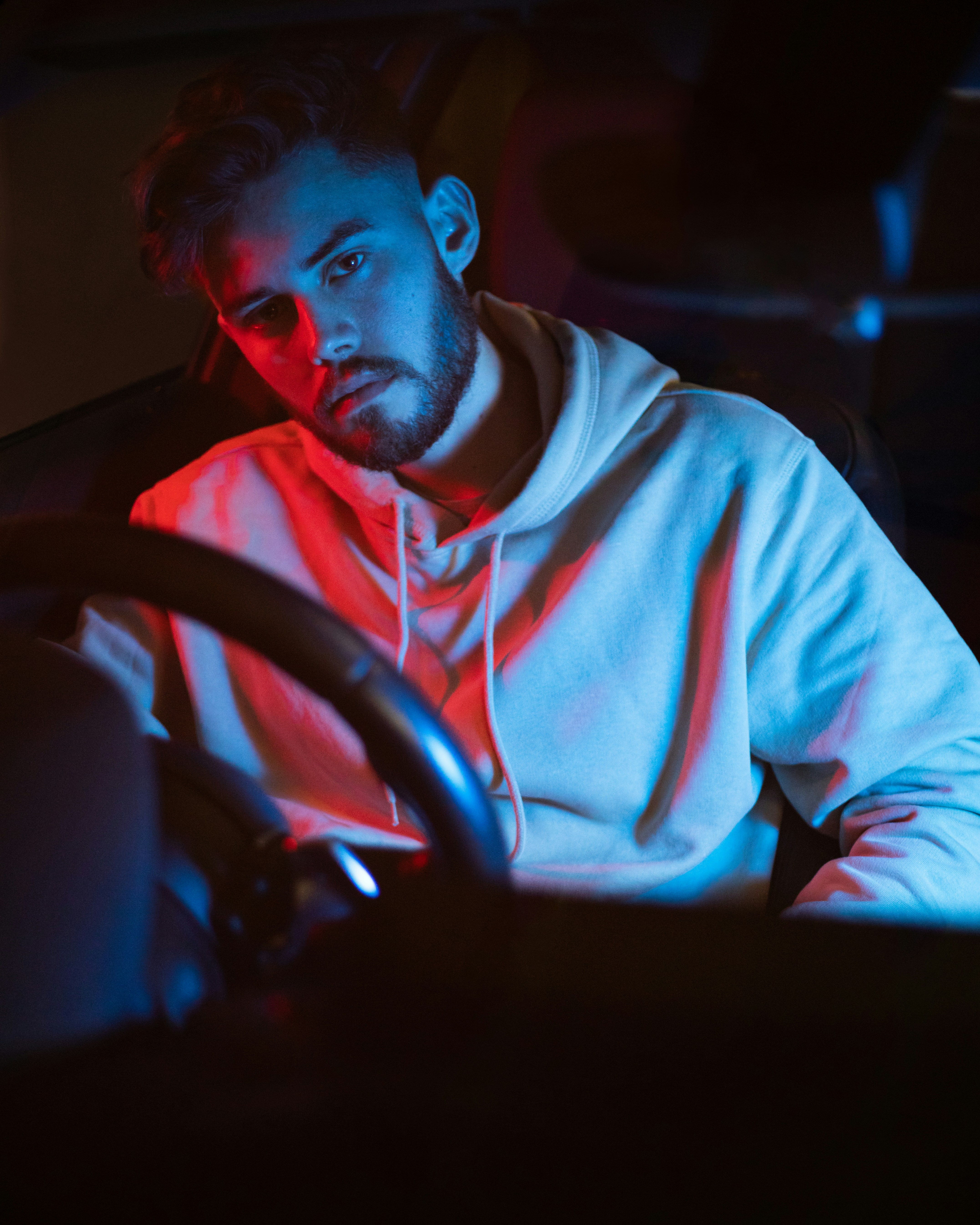 Photograph of a man in a hoodie inside a car at night, illuminated by red and blue neon, gazing toward the camera.