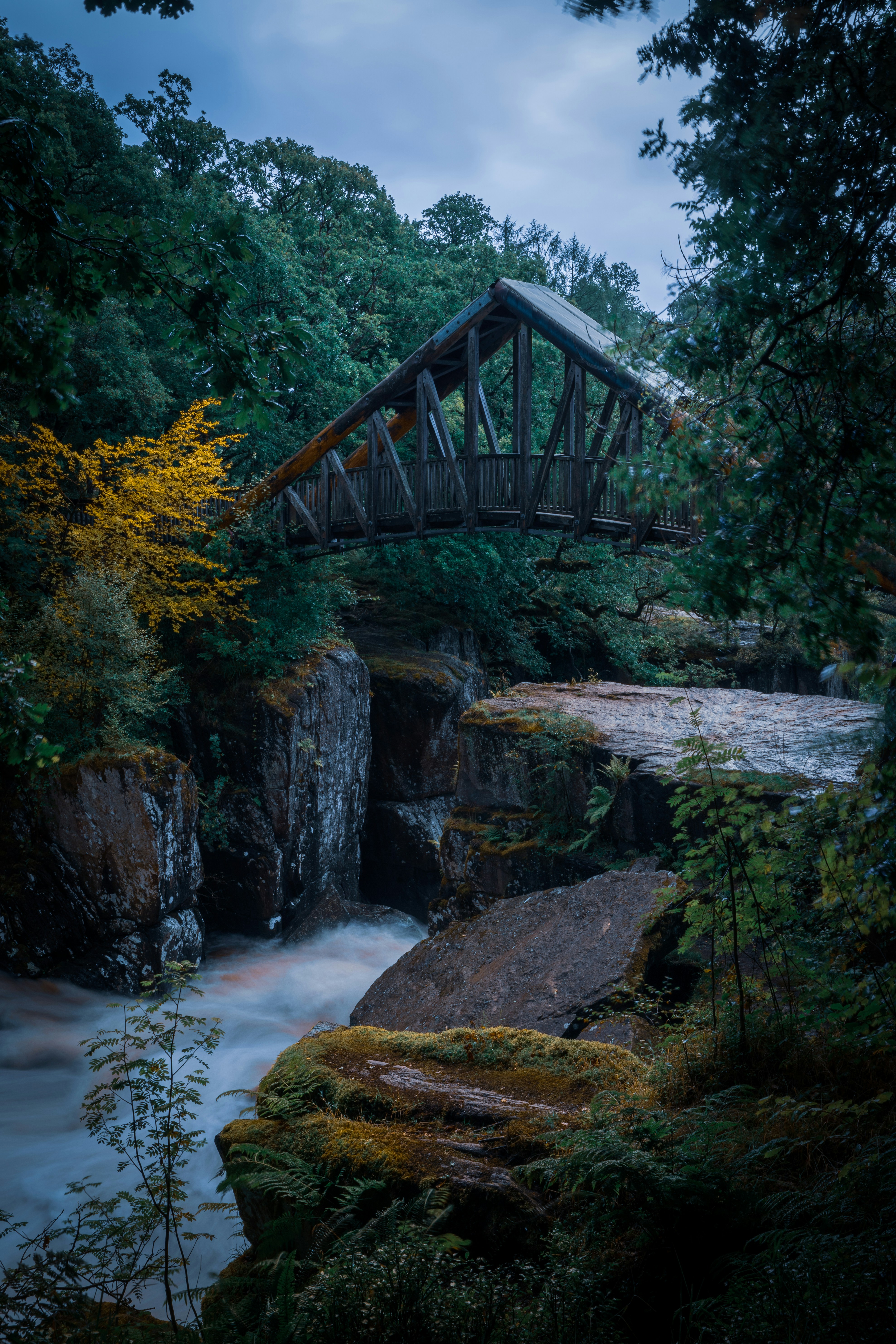 A wooden bridge arches gracefully over a serene river, surrounded by lush greenery and autumn foliage. The gentle flow of water adds to the peaceful atmosphere.
