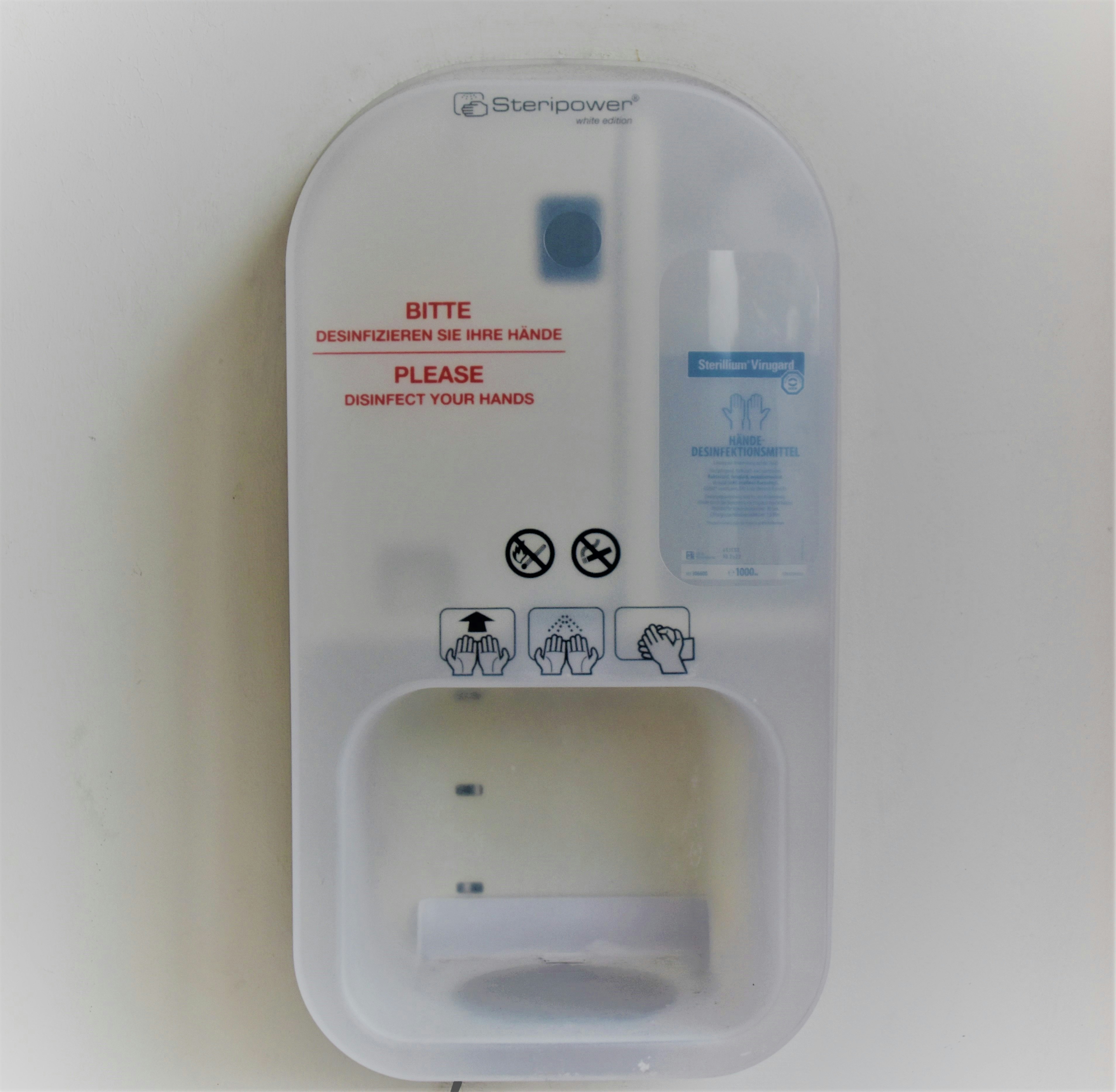 Wall-mounted hand sanitizer dispenser with clear instructions in German, promoting hygiene practices. Contains a visible bottle of sanitizer and graphical handwashing instructions.