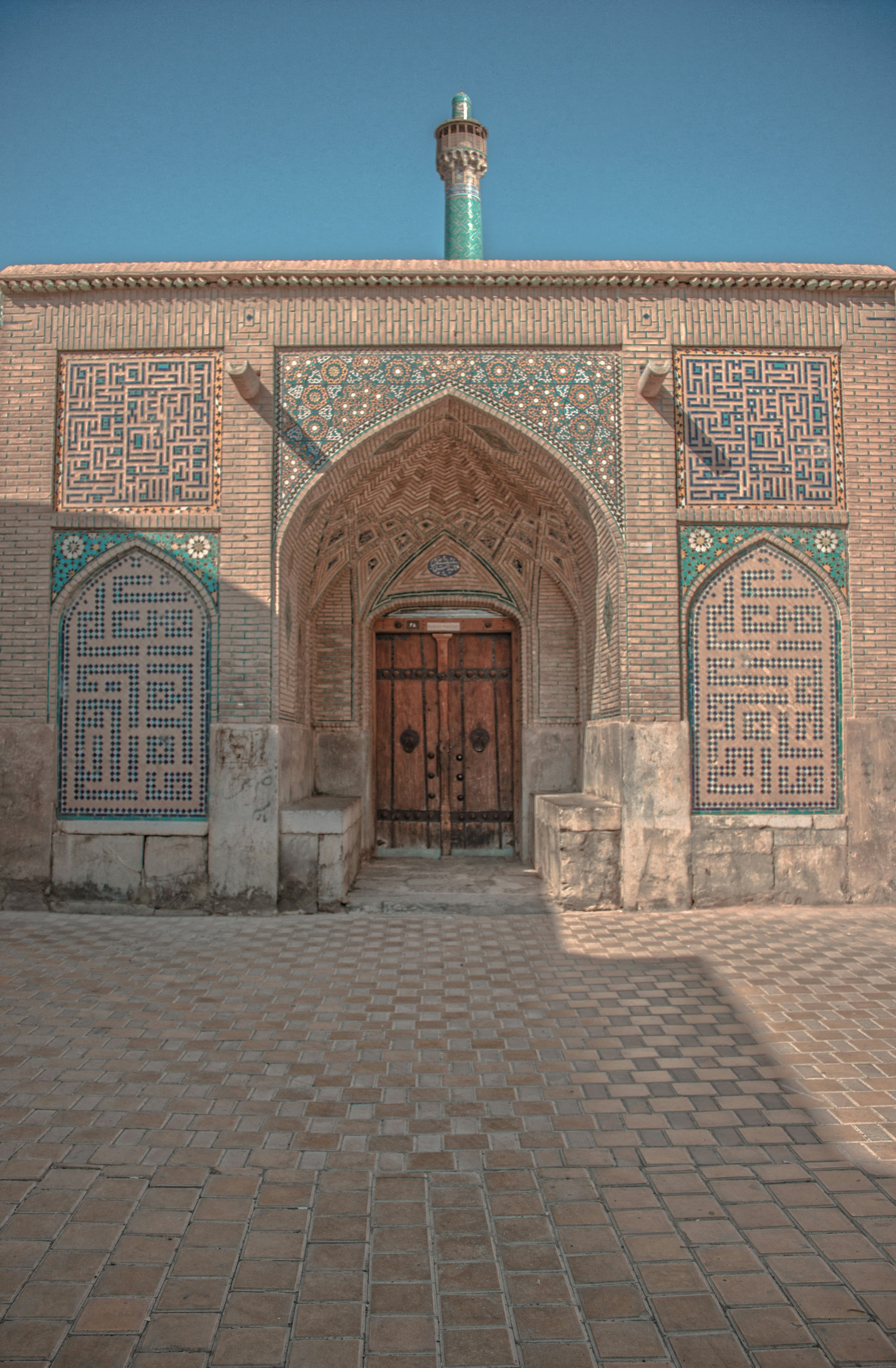 Intricate brick archway adorned with colorful tile work, leading to a wooden door, showcasing traditional craftsmanship. The structure stands against a clear blue sky.