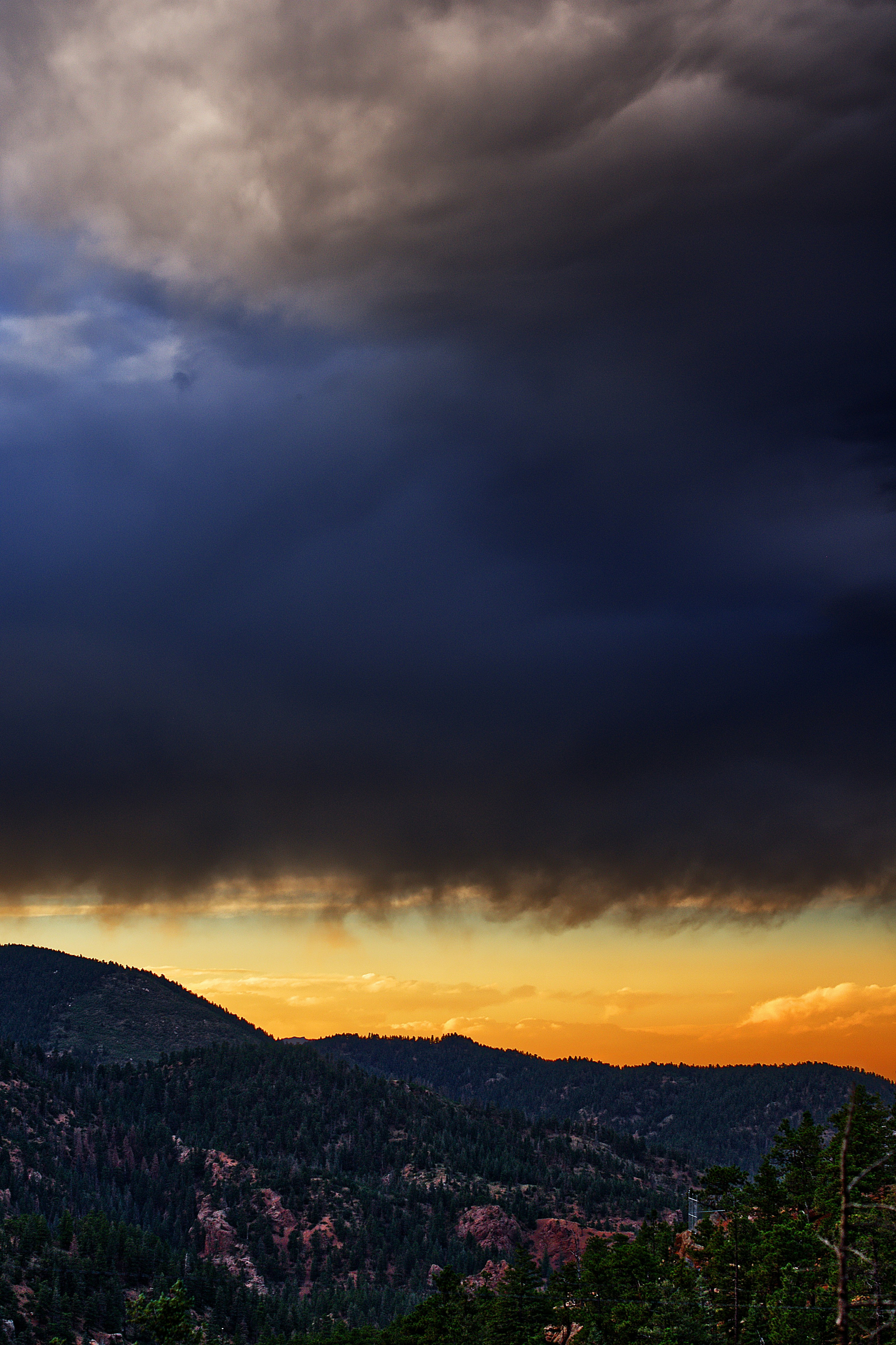 Dark clouds loom over a mountainous landscape, with a warm glow of sunset illuminating the horizon. The scene captures the transition from day to night.