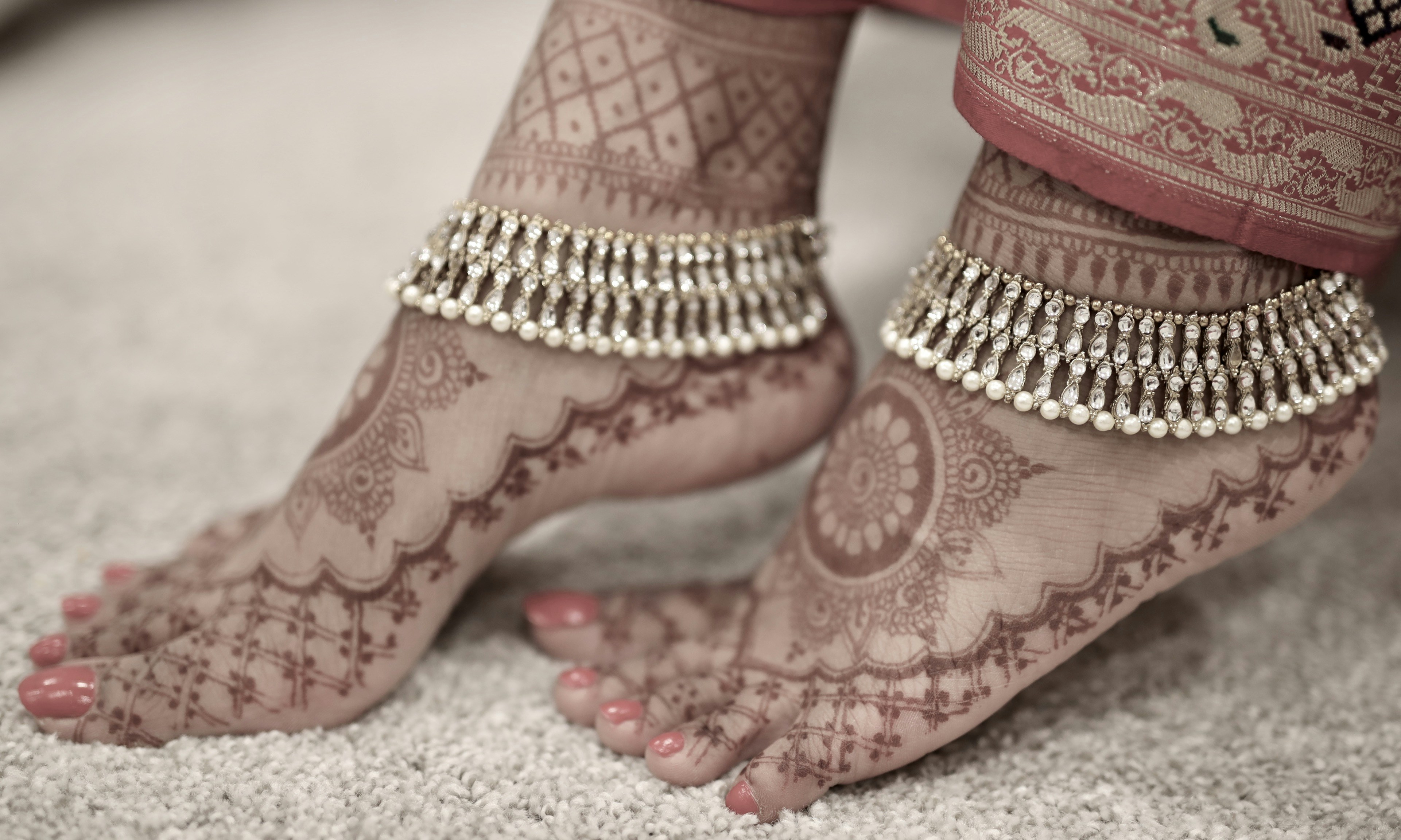 Intricately designed henna patterns on feet adorned with silver anklets, resting on a soft surface. The cultural significance of the designs highlights a celebration of tradition.