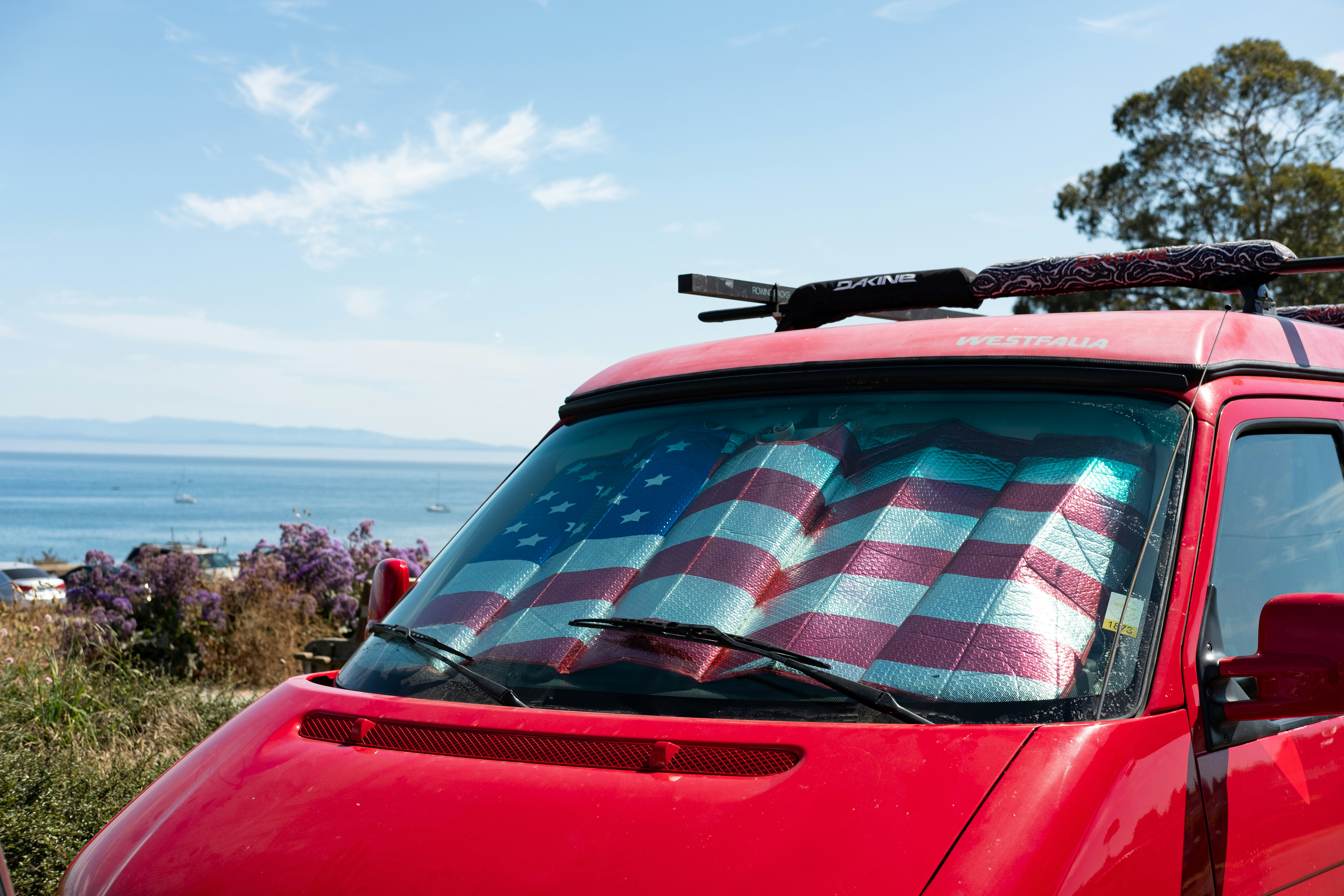 Red vehicle with an American flag sunshade parked near a scenic coastal view, surrounded by lush greenery and vibrant flowers.