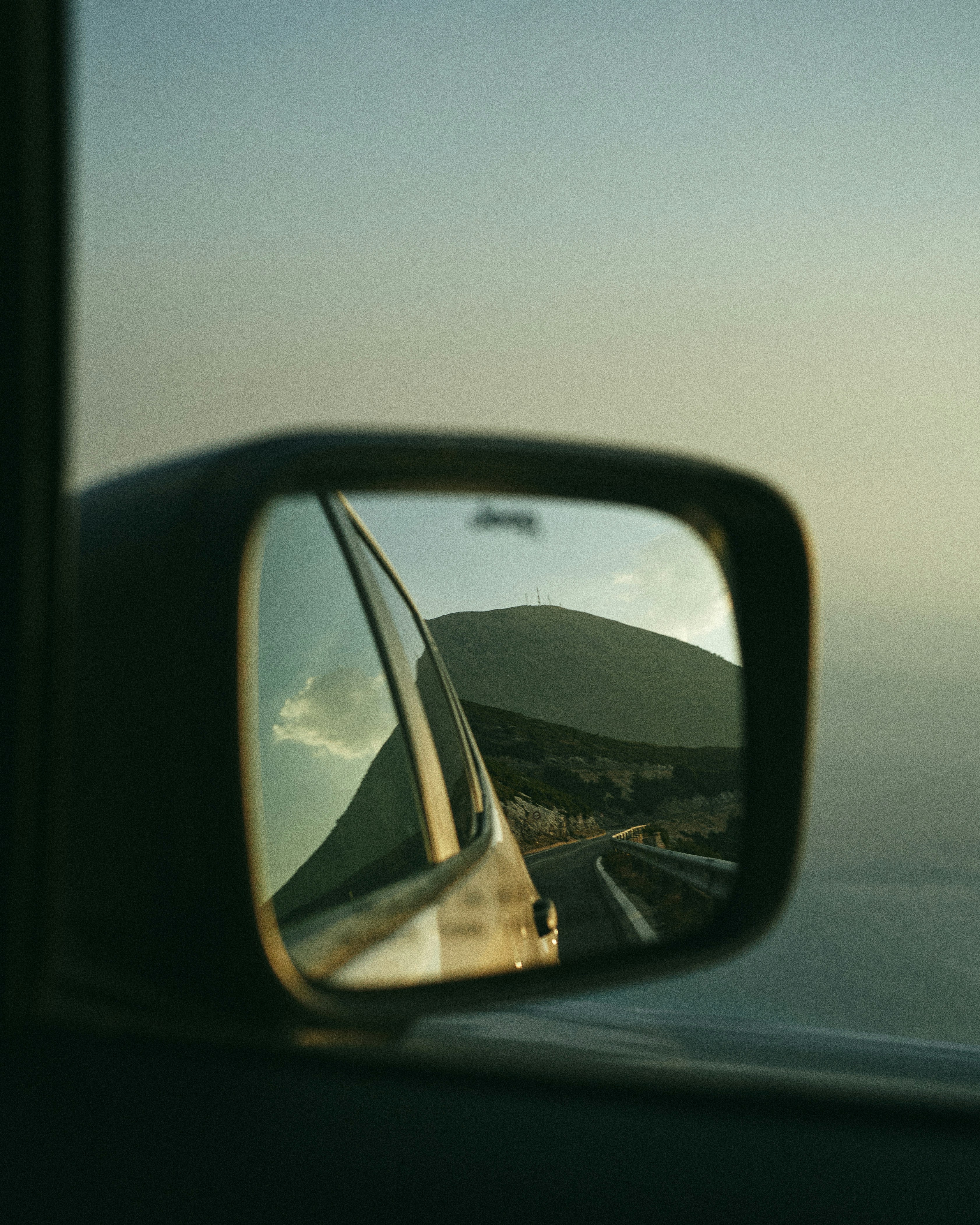 Side-view mirror frames a coastal road winding up a hillside at sunset, with the sea visible in the distance.