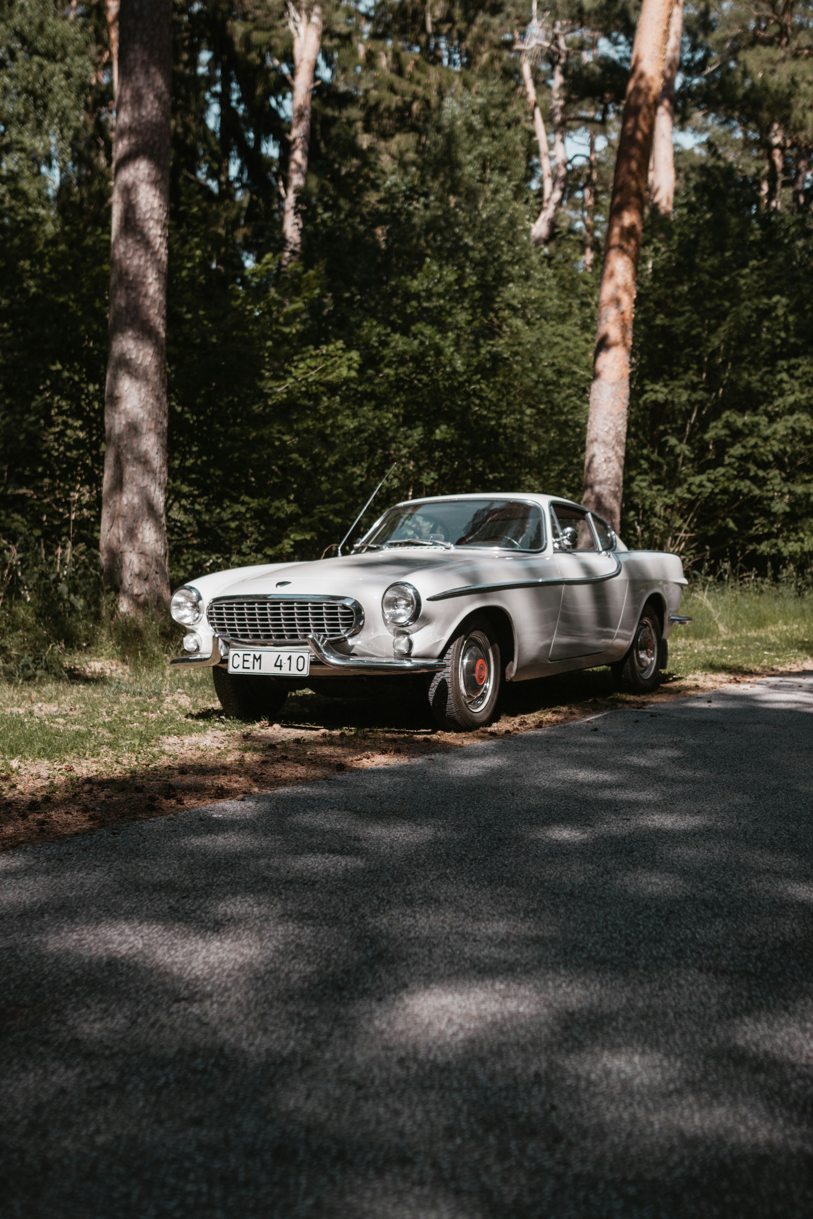 Classic car parked beside a winding road, surrounded by lush greenery and tall trees. The scene captures a moment of tranquility in nature.