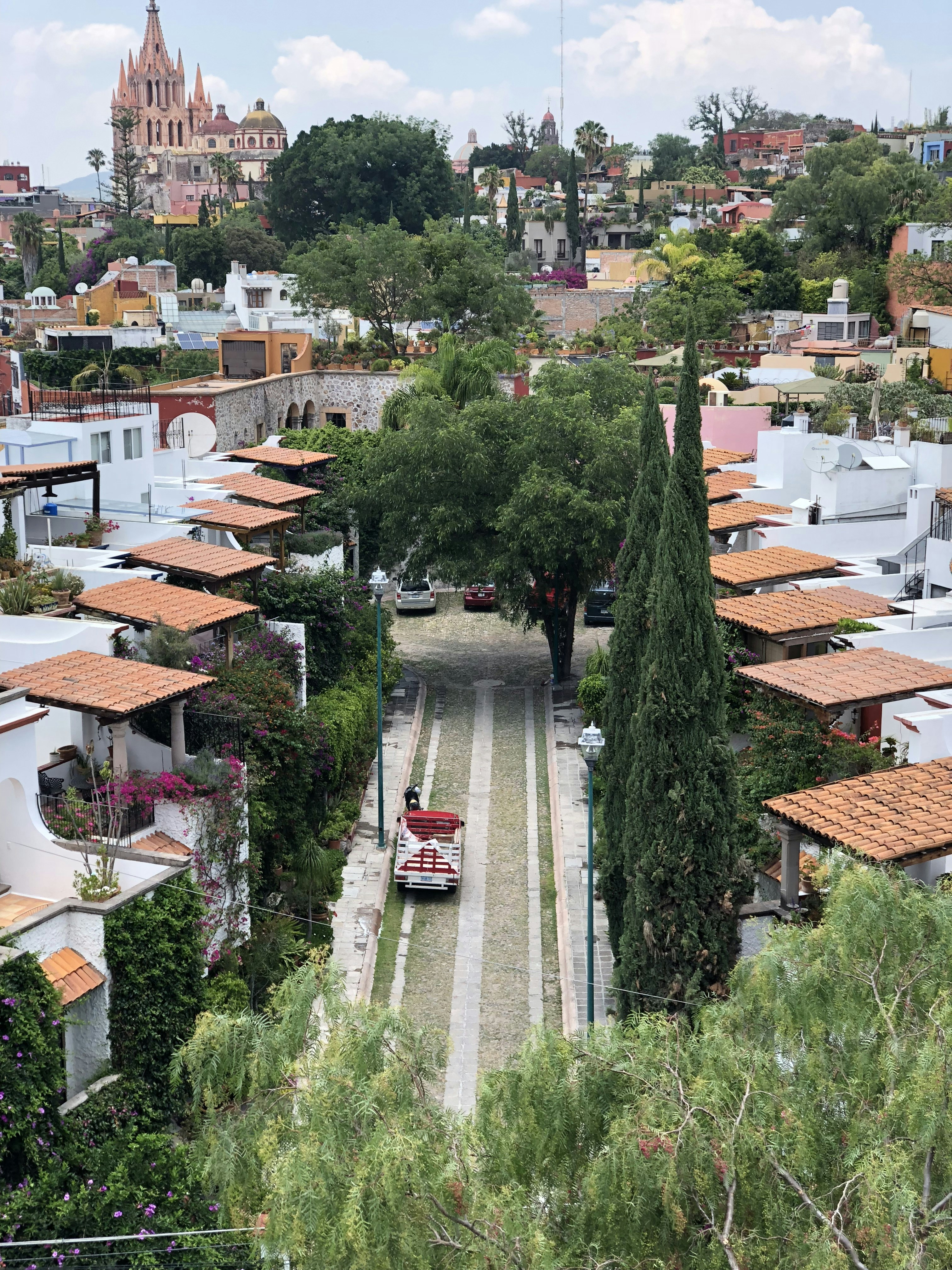 Charming pathway lined with vibrant houses and lush greenery, leading to a distant church steeple under a cloudy sky.