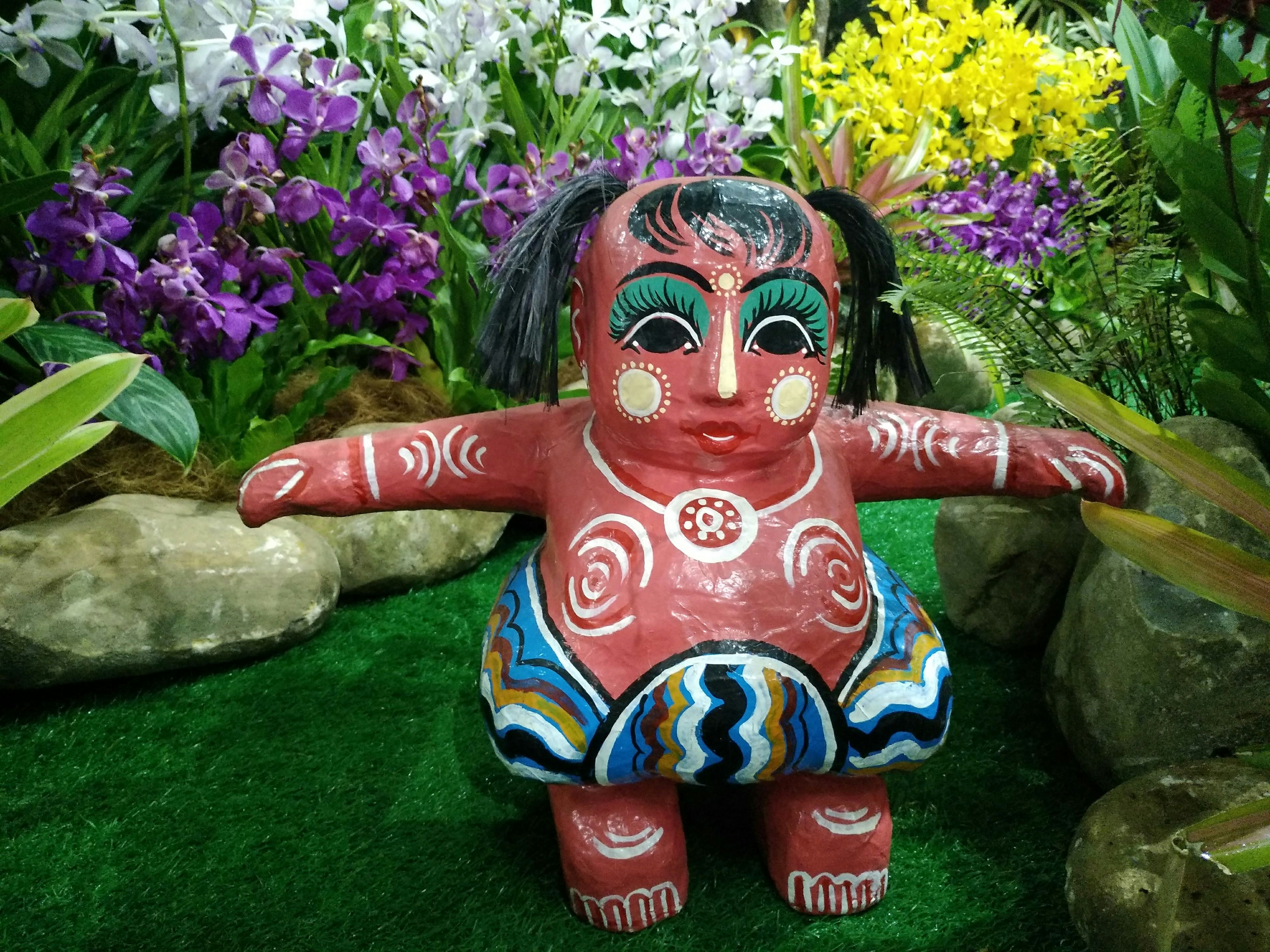 Colorful figurine with intricate designs surrounded by vivid flowers and greenery.