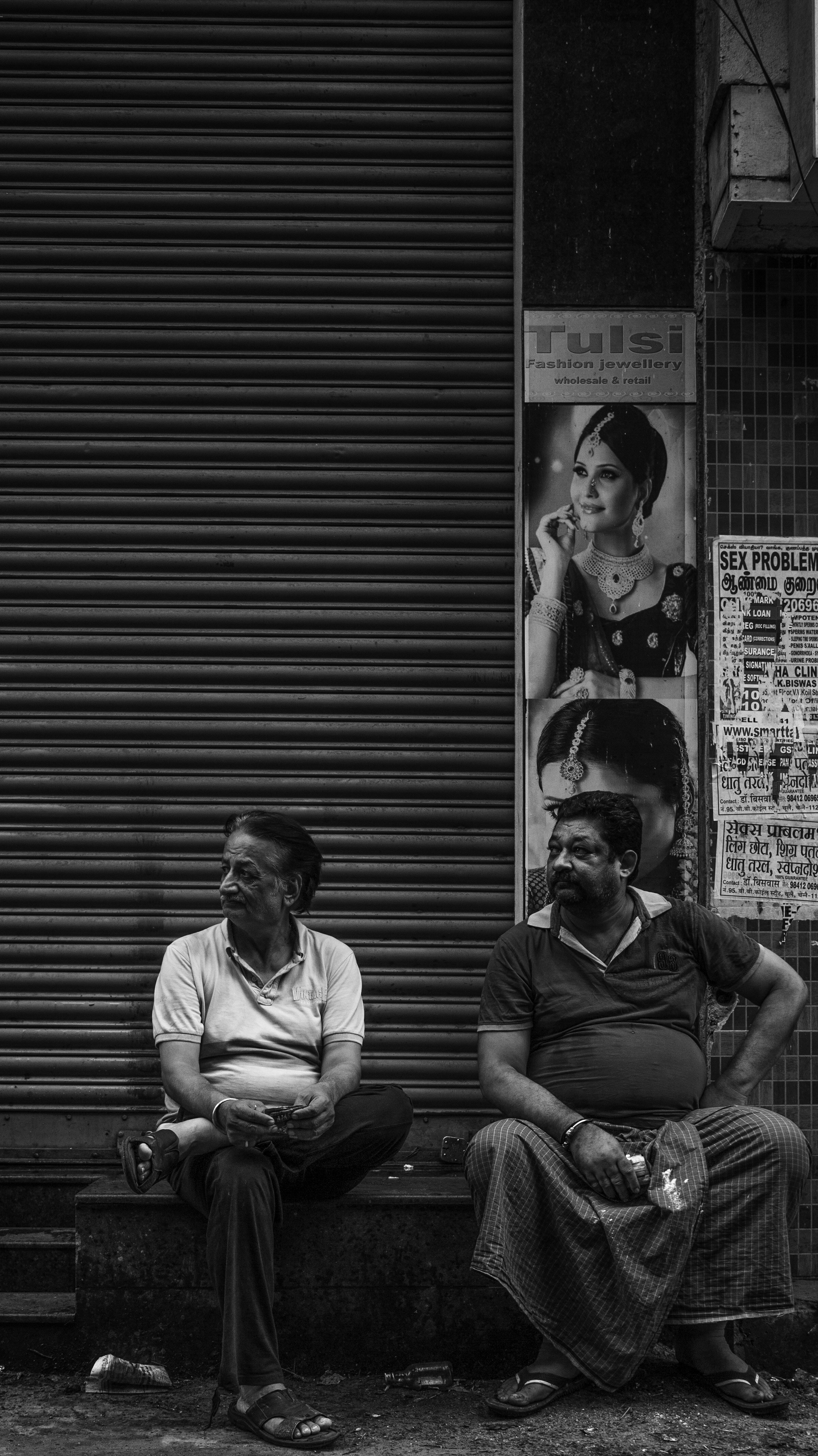Two men sitting on a street curb, lost in thought, with a fashion advertisement on the wall behind them.