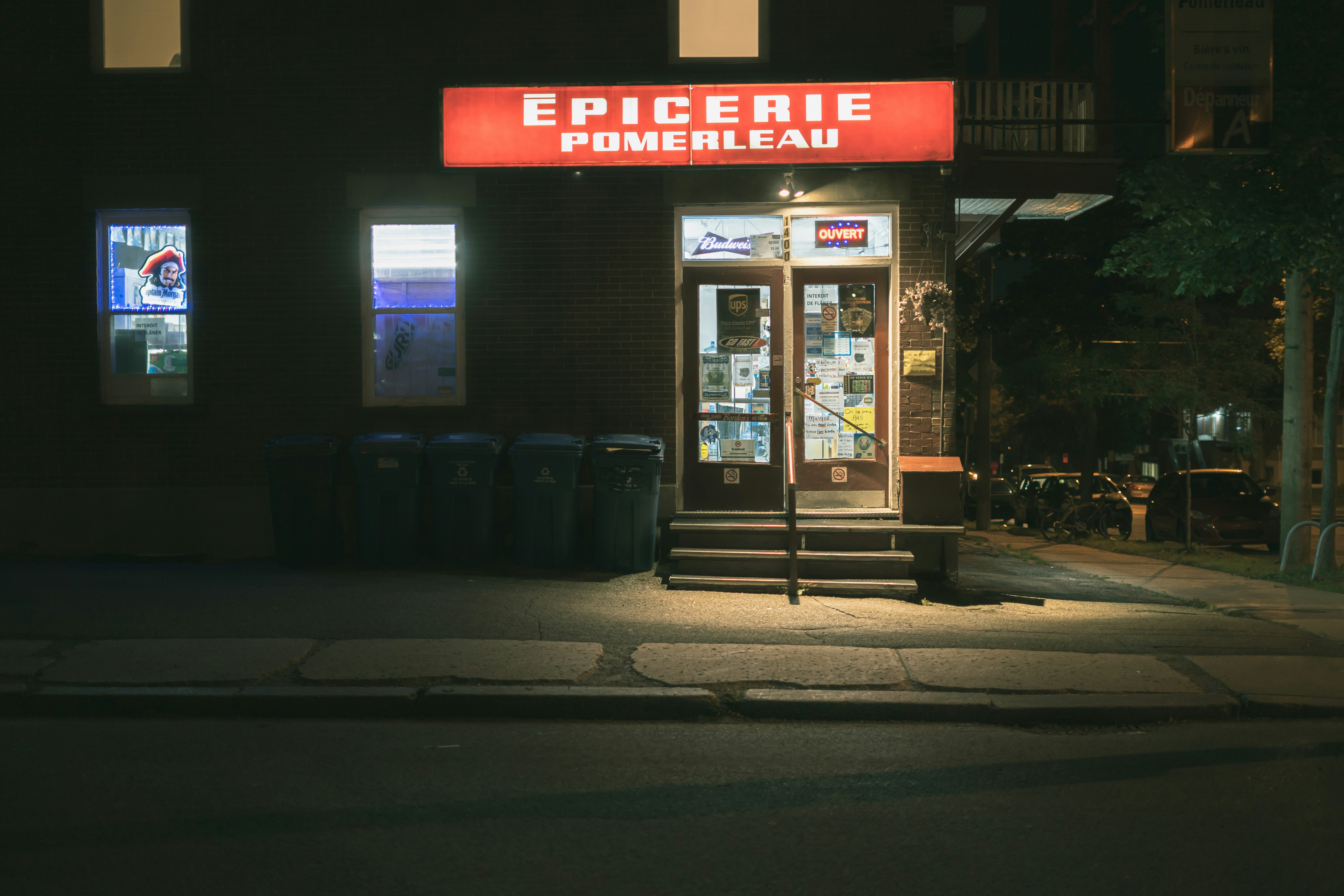 This image captures a quaint local store under the soft glow of night lights, with a prominent red sign that reads 'Épicerie Pomerleau'. The scene is bathed in warm, ambient lighting that contrasts with the cool blues of nearby windows, creating a visually striking composition. The tranquil atmosphere, enhanced by the shadows and streetlamp illumination, evokes a sense of calm and nostalgia, making the storefront the focal point against the darkened street.