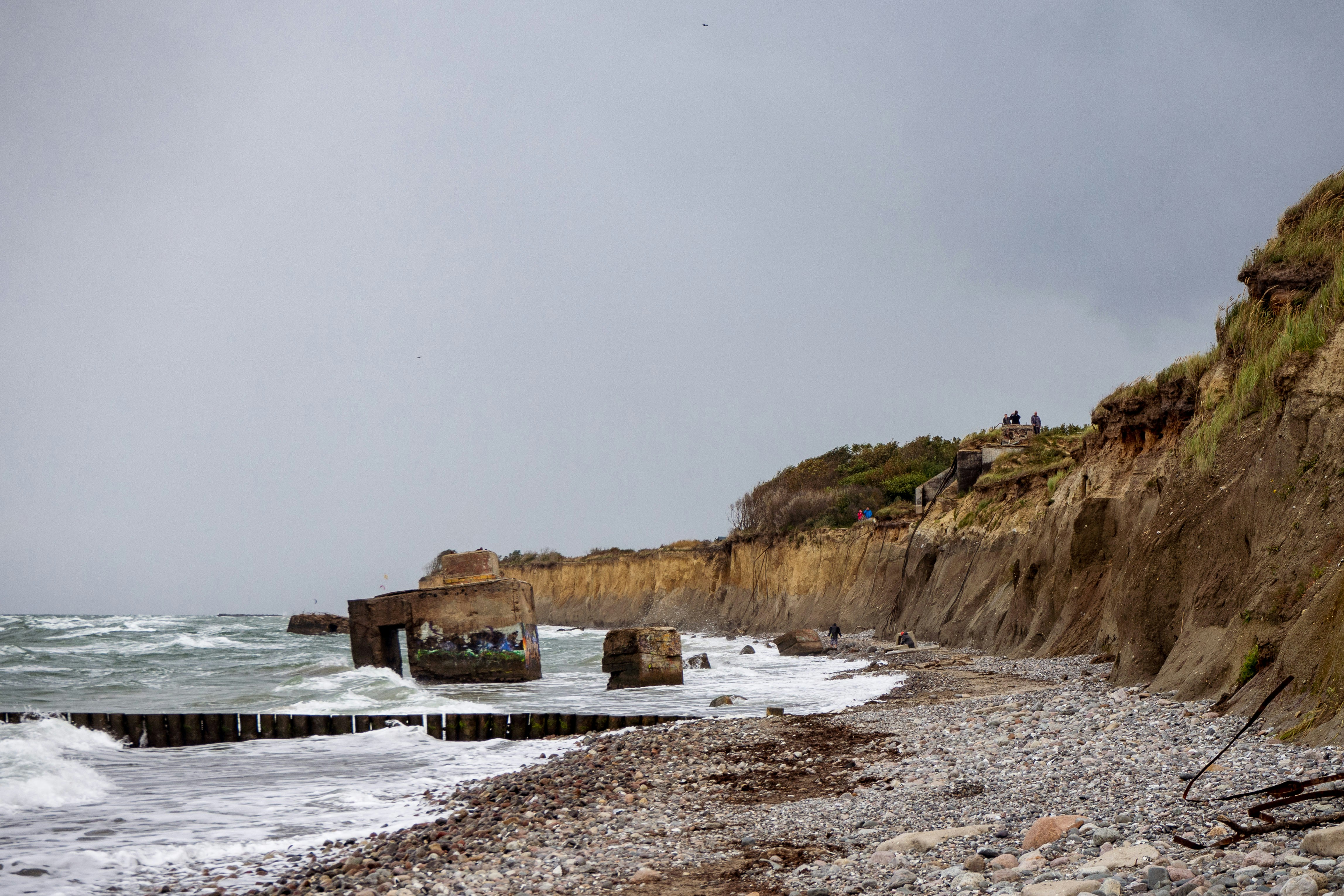 Weathered remnants of a structure stand against the crashing waves, framed by a rugged coastline and overcast skies.