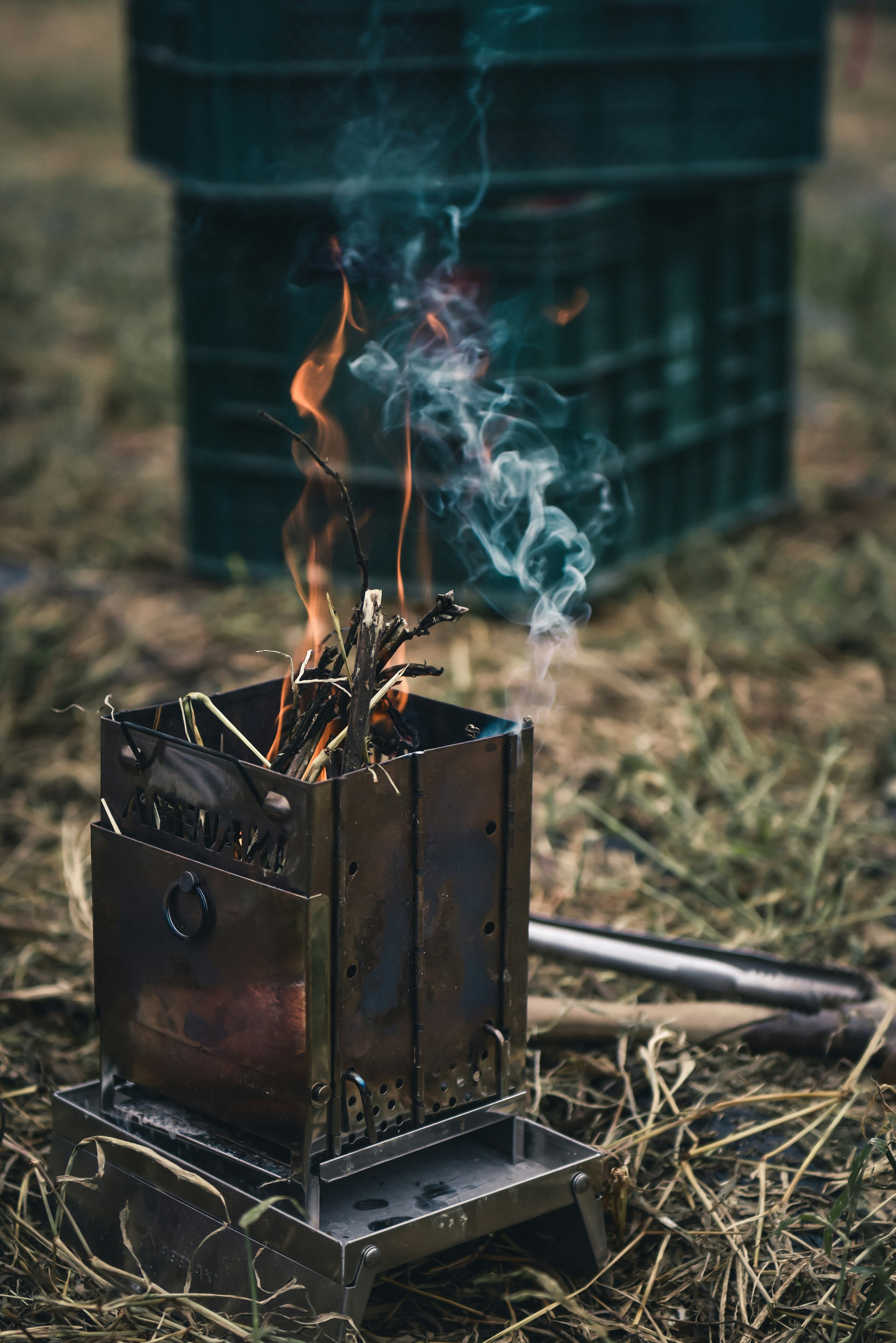 Portable camping stove ignited with twigs, releasing flames and smoke amidst a grassy backdrop.