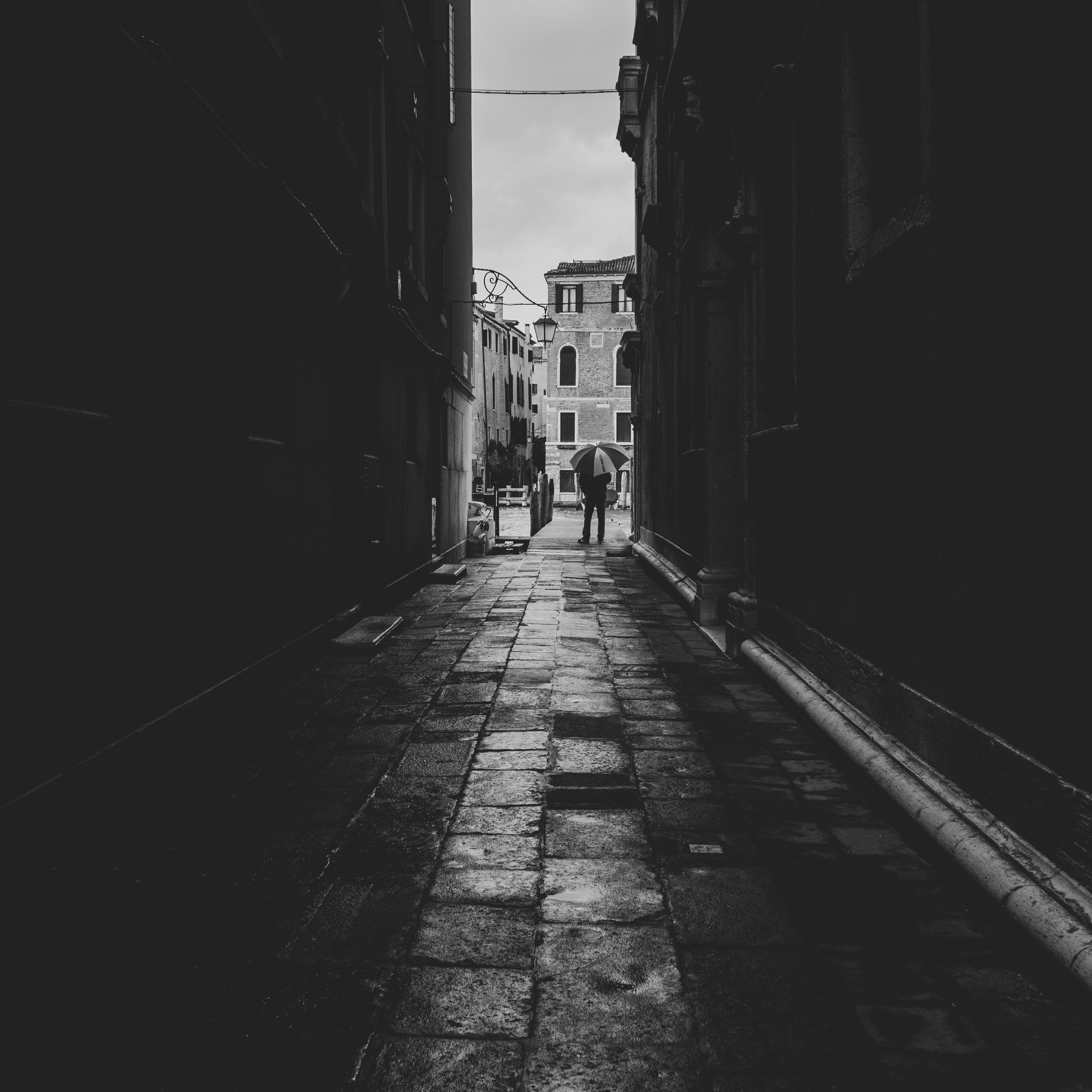 Two figures walking through a narrow, dimly lit alley in Venice, surrounded by shadowy buildings. The wet cobblestones reflect the muted light.
