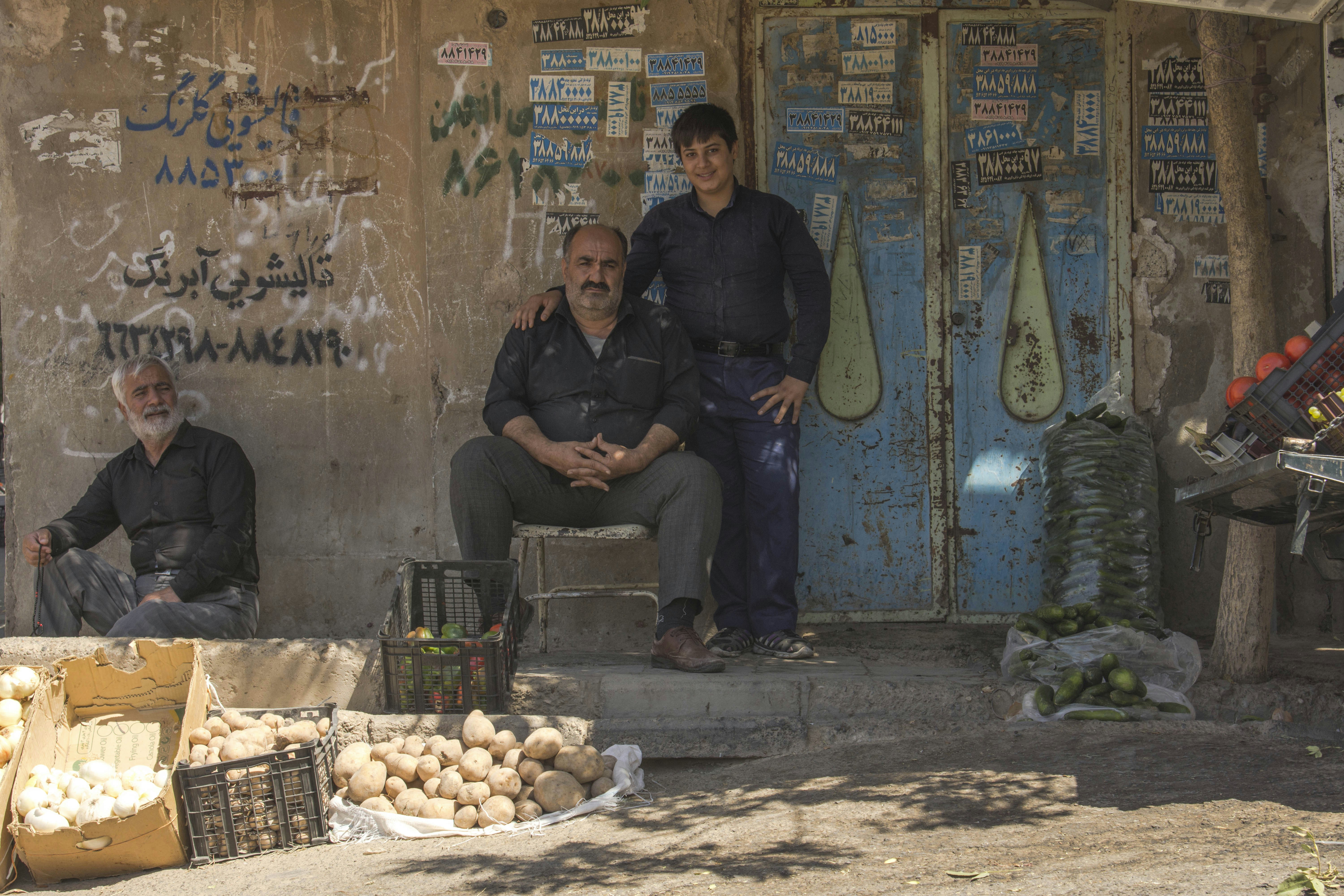 Two vendors pose in front of their shop, surrounded by fresh produce and handwritten signs. The scene captures the essence of local commerce and community spirit.
