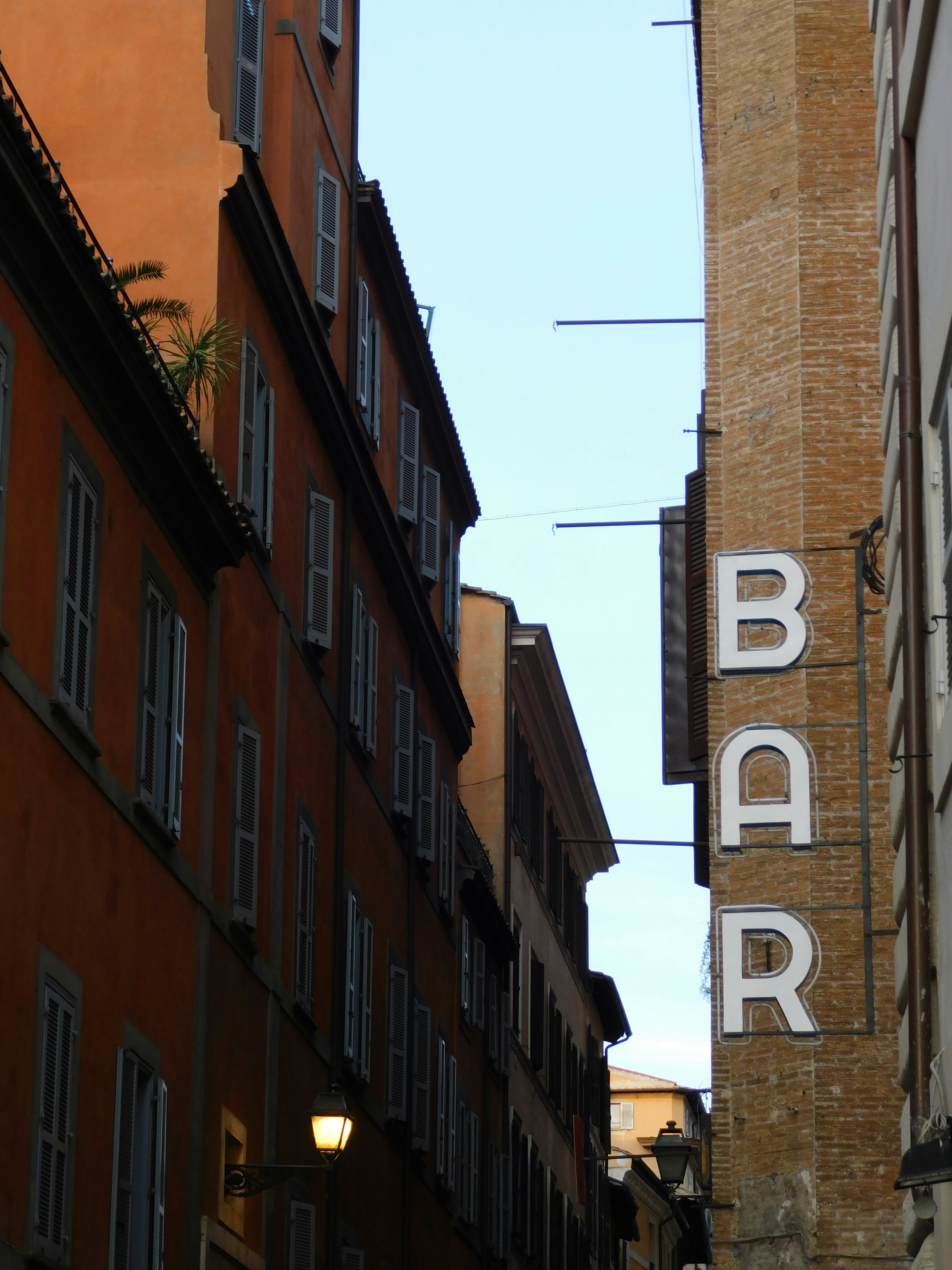 Narrow street flanked by colorful buildings, featuring a prominent bar sign against a clear sky. Street lamps add a warm glow to the scene.