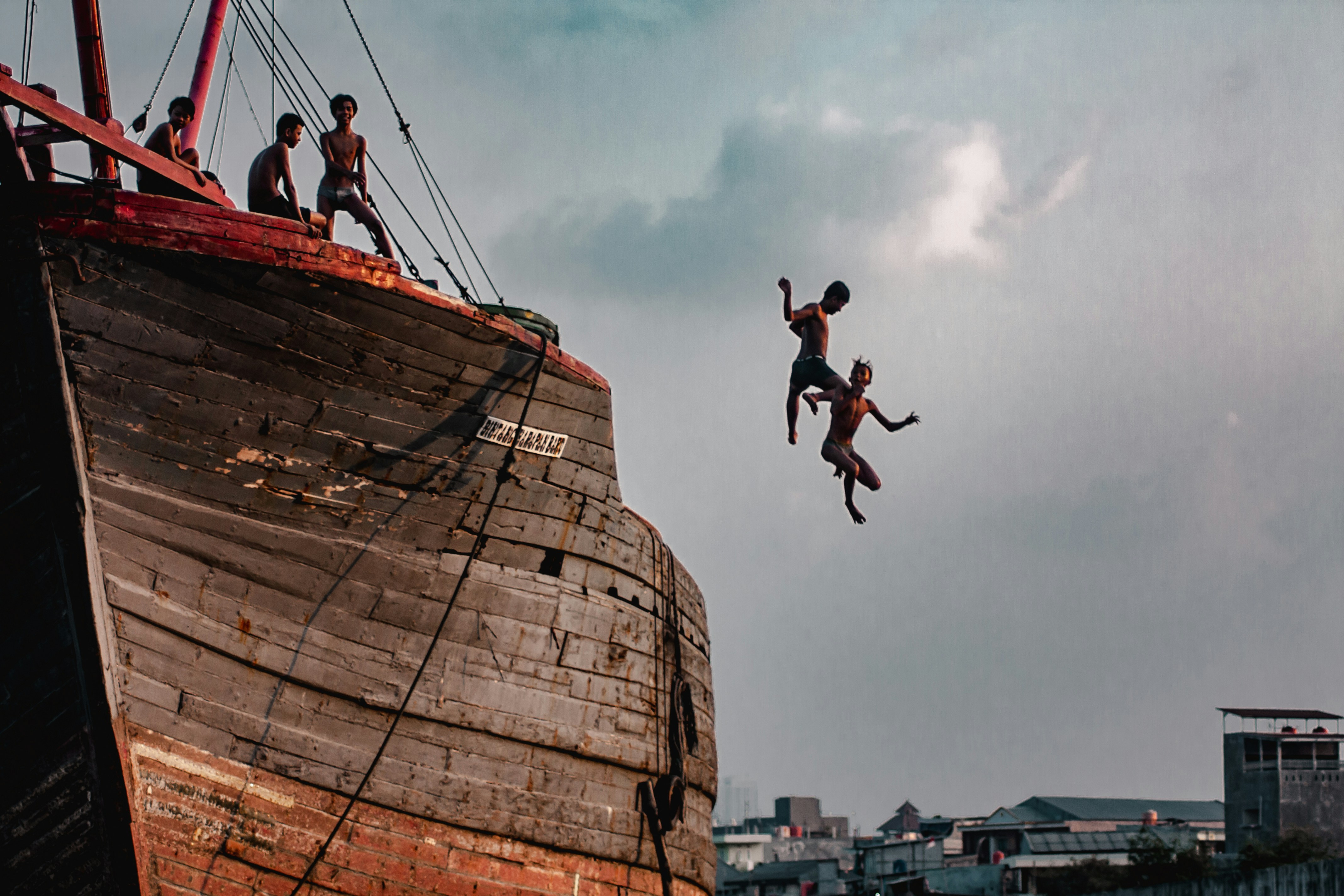 Two people mid-air after jumping off a large wooden ship into the water below, with an overcast sky in the background.