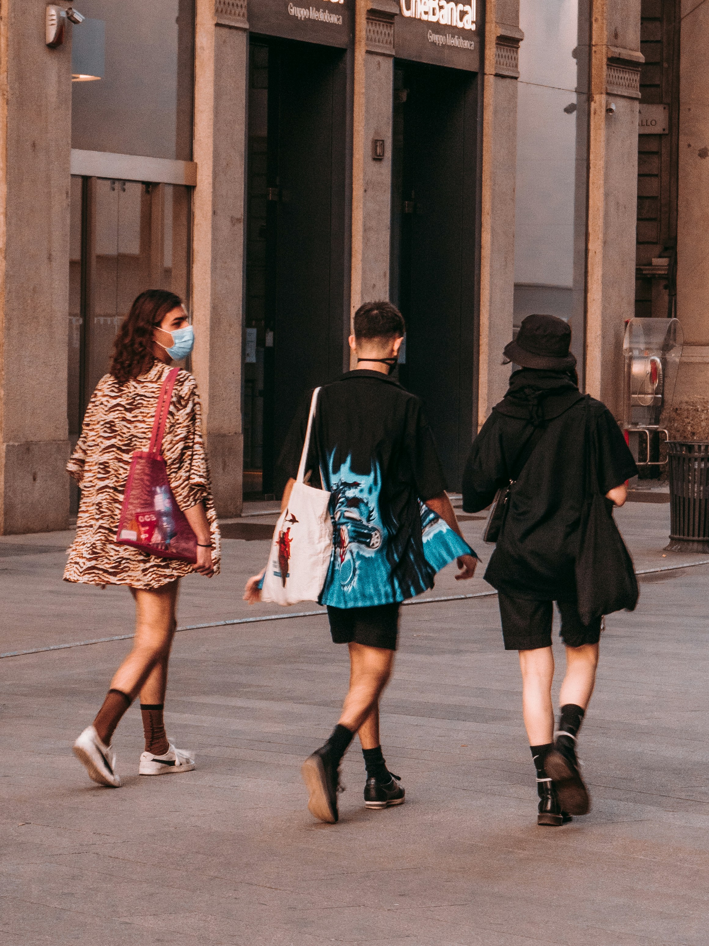 Three individuals stroll through a city street, showcasing eclectic fashion styles and accessories. The scene captures a blend of contemporary urban culture.