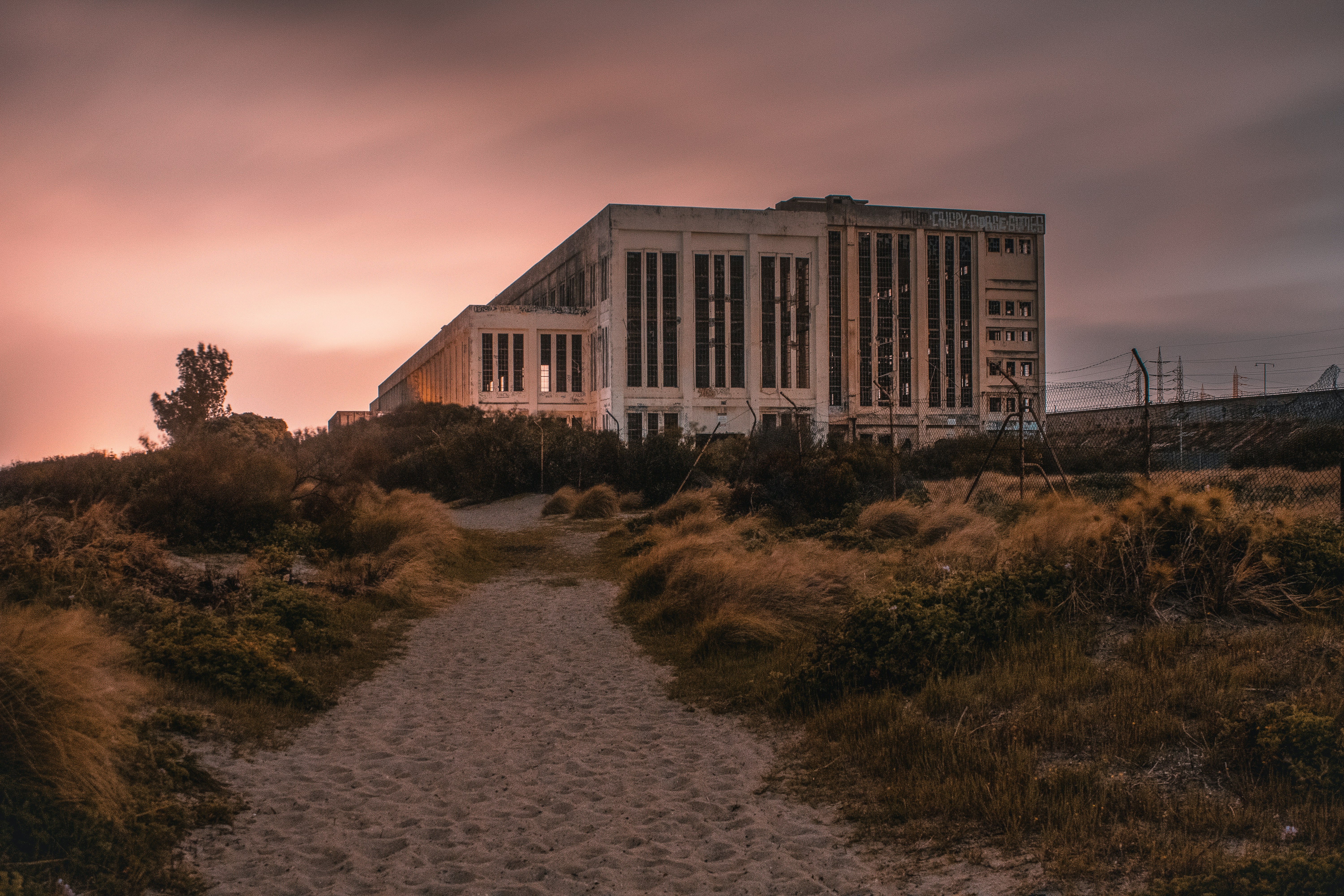 Dune path leads to a stark, modern building at sunset with warm, dusky sky and soft foreground contrast. The scene emphasizes isolation of the structure against natural brush.