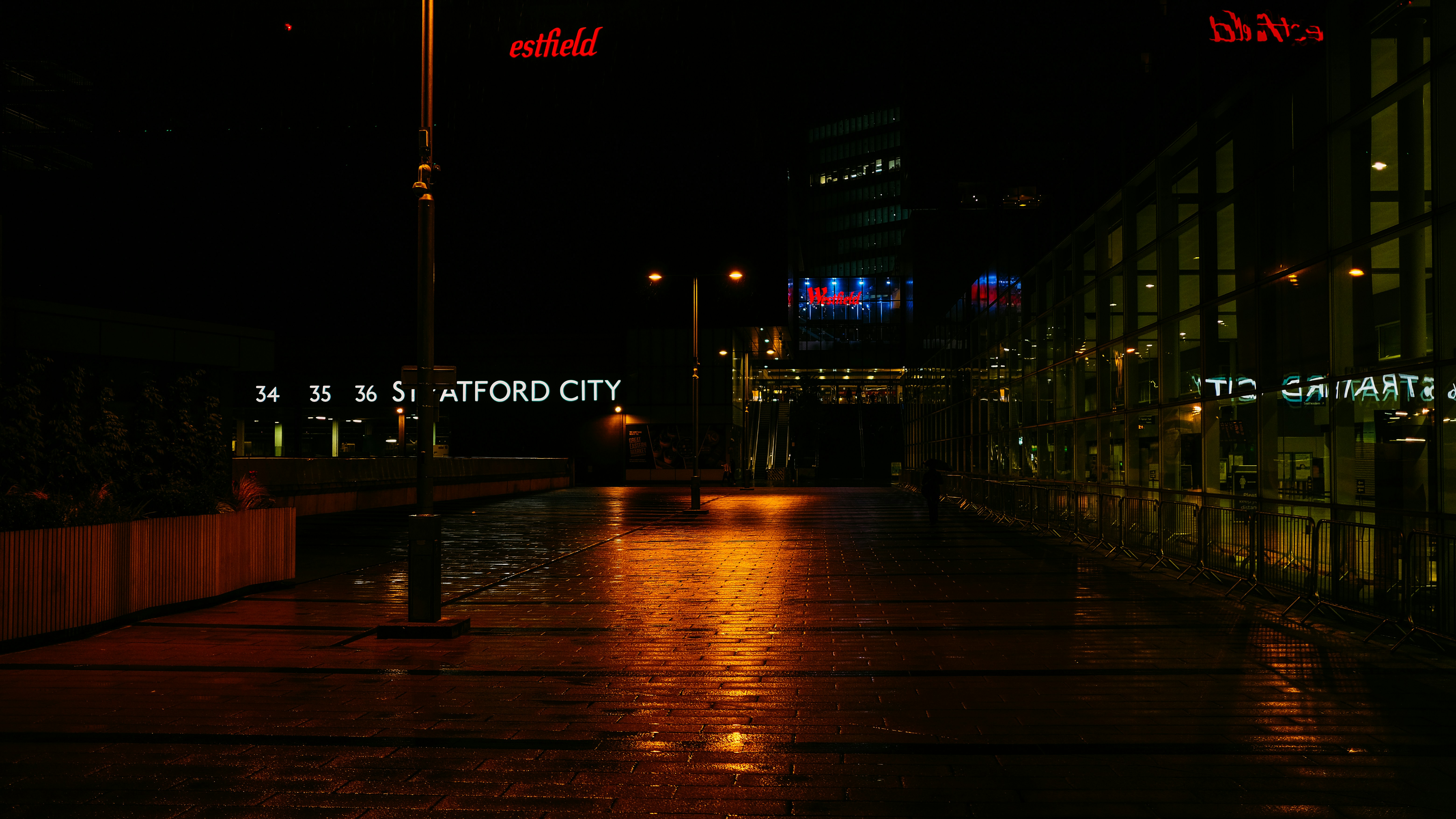 Wet pavement reflecting city lights in Stratford City at night, with illuminated signage in the background.