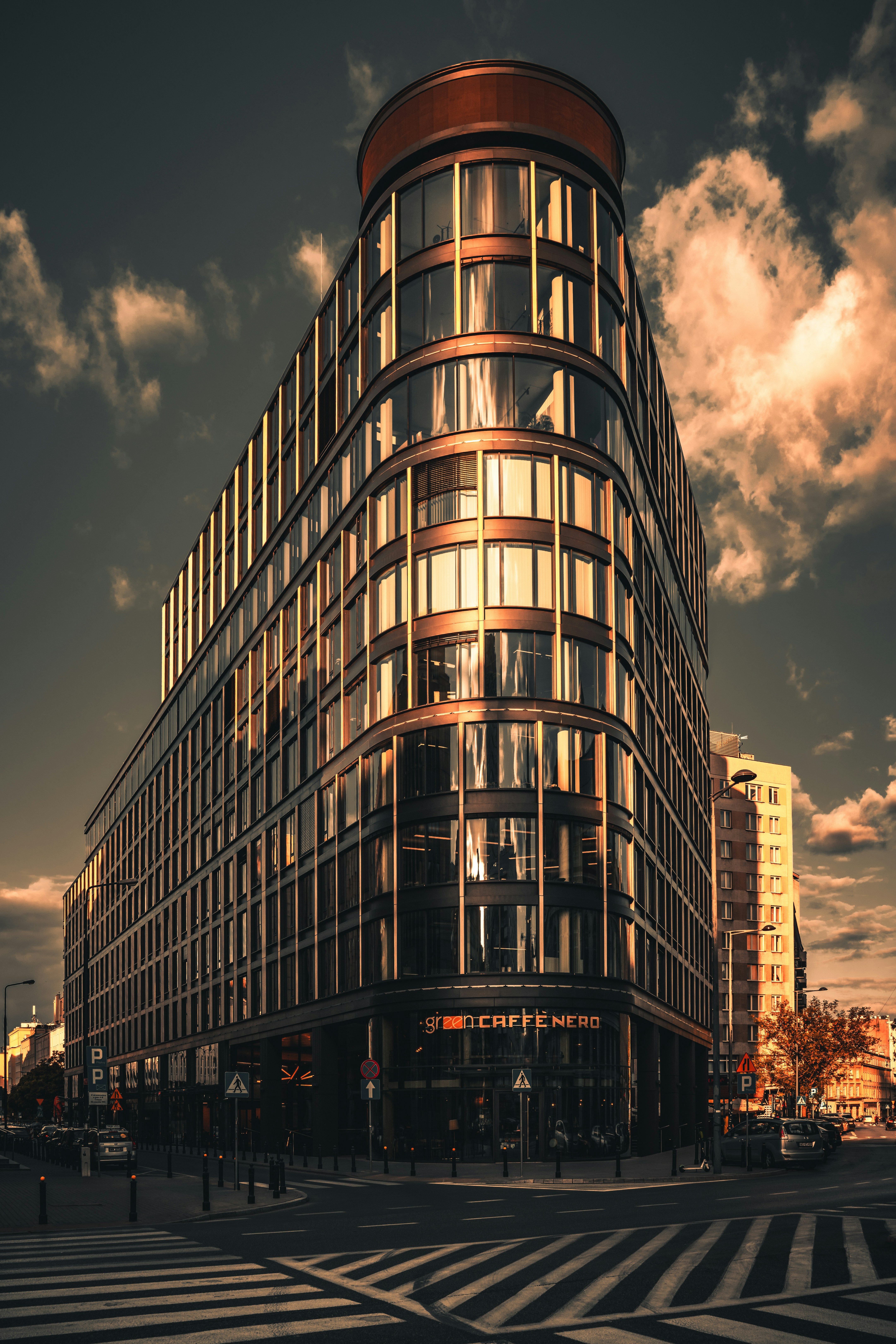 Contemporary building with sleek glass and metal design reflecting the evening light, showcasing urban architecture. The scene captures a bustling street corner with traffic and pedestrians.