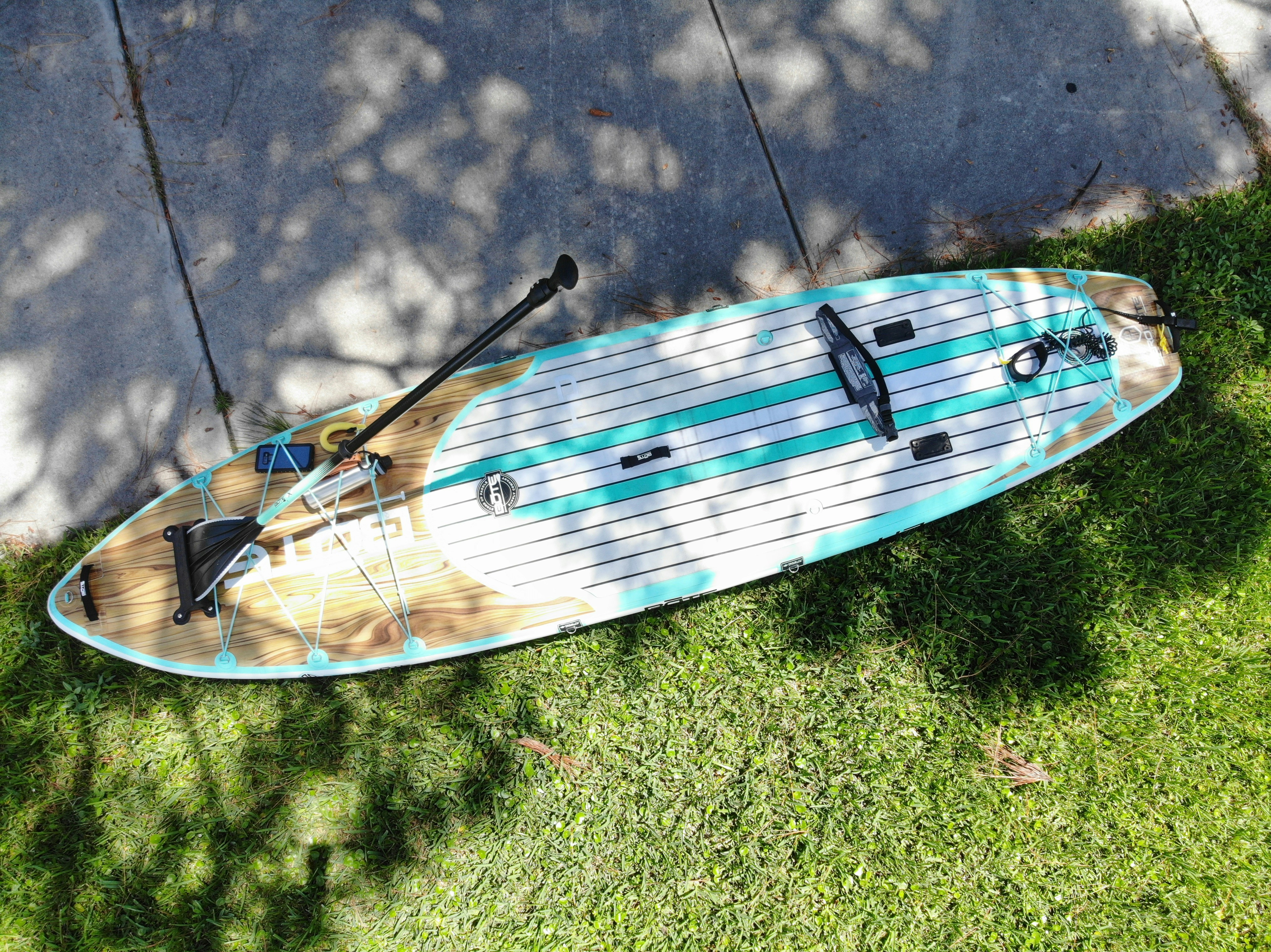 Striped stand-up paddleboard resting on sunlit grass beneath dappled shade