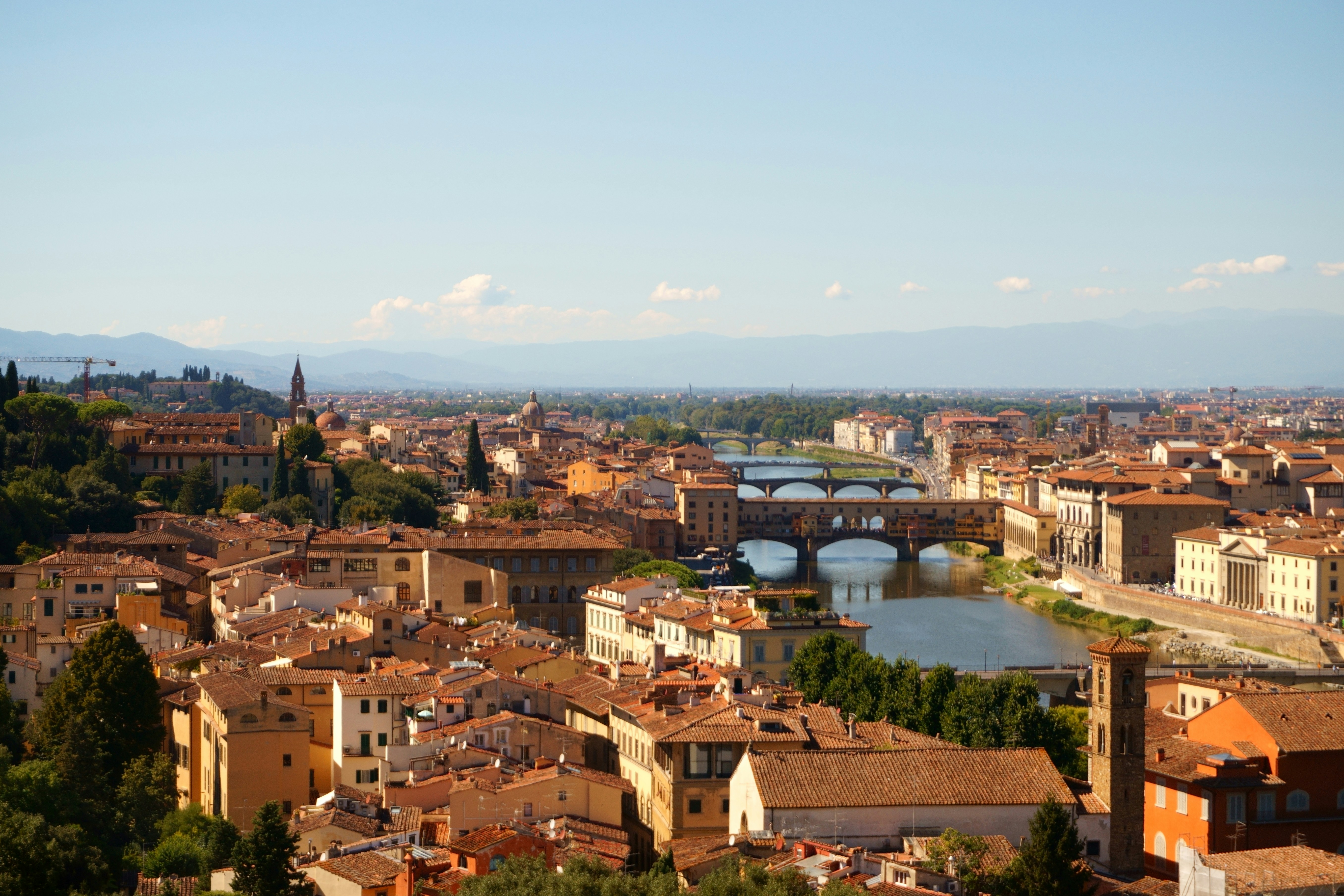 Panoramic view of Florence showcasing the Arno River and iconic bridges amidst terracotta-roofed buildings under a clear blue sky.