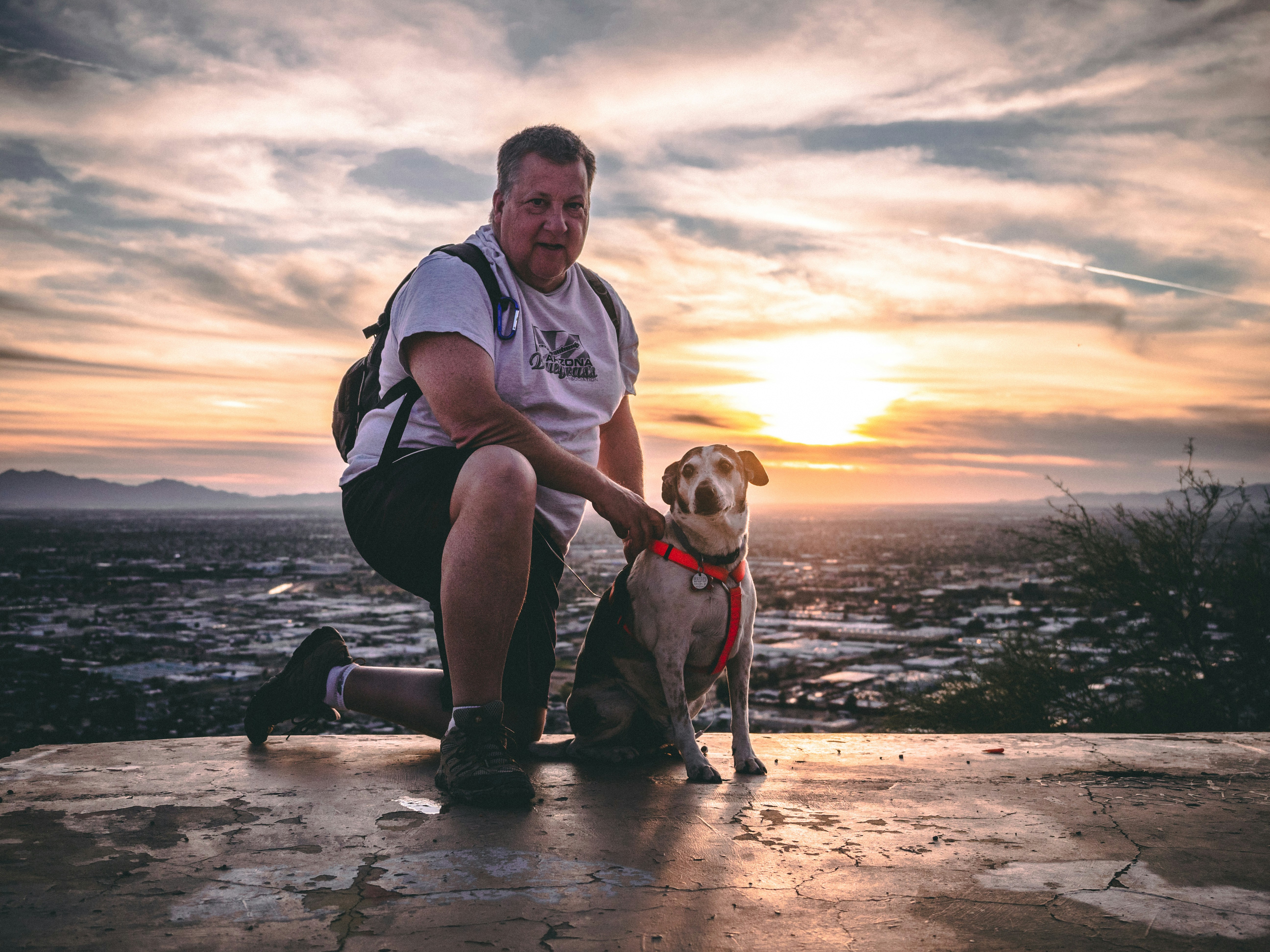 A man kneels beside his dog on a rocky outcrop, with a vibrant sunset casting warm hues over the landscape.