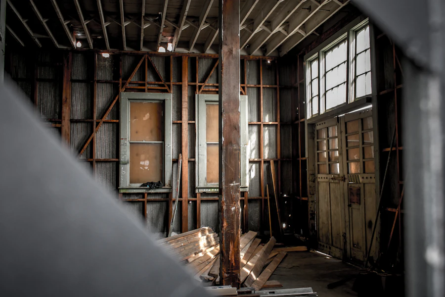 Empty industrial room with exposed studs, corrugated metal walls, and boarded windows lit by soft daylight. A central support post and stacked lumber add to the unfinished, abandoned feel.