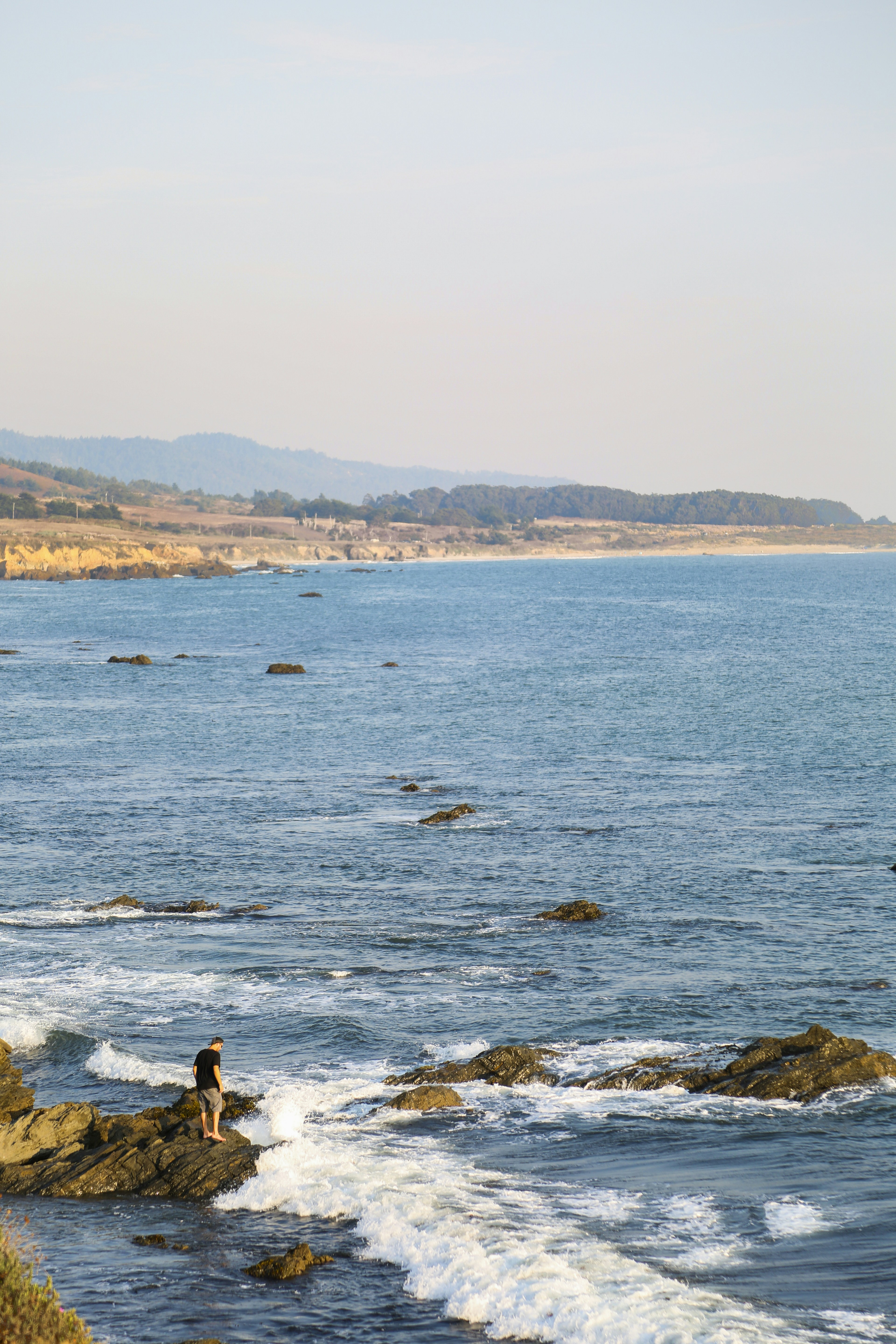 A lone figure stands on rocky outcrops by the ocean, gazing at the waves as the sun casts a warm glow over the landscape.