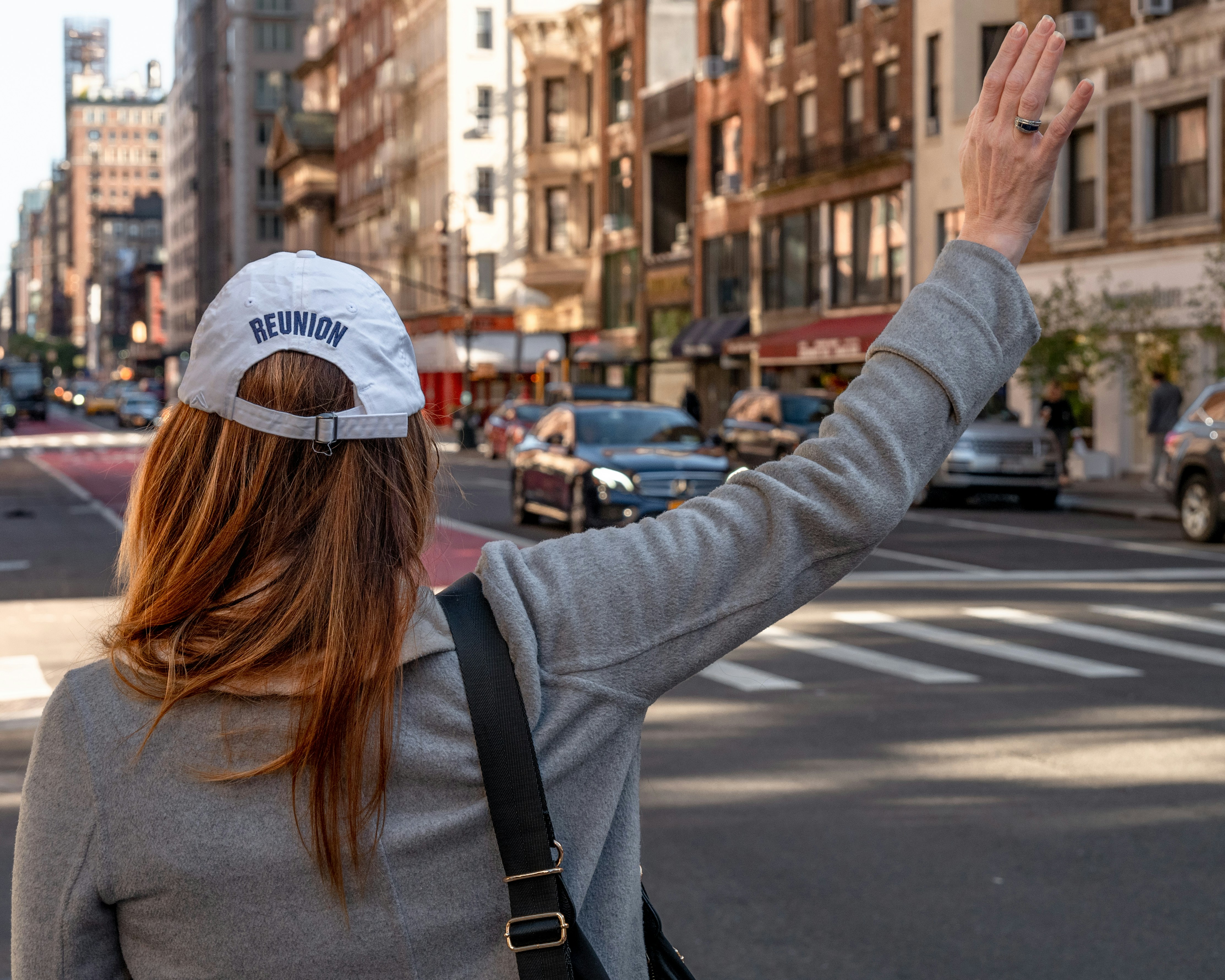 Woman in a gray coat and cap with 'REUNION' raised hand hailing a taxi on a bustling city street.