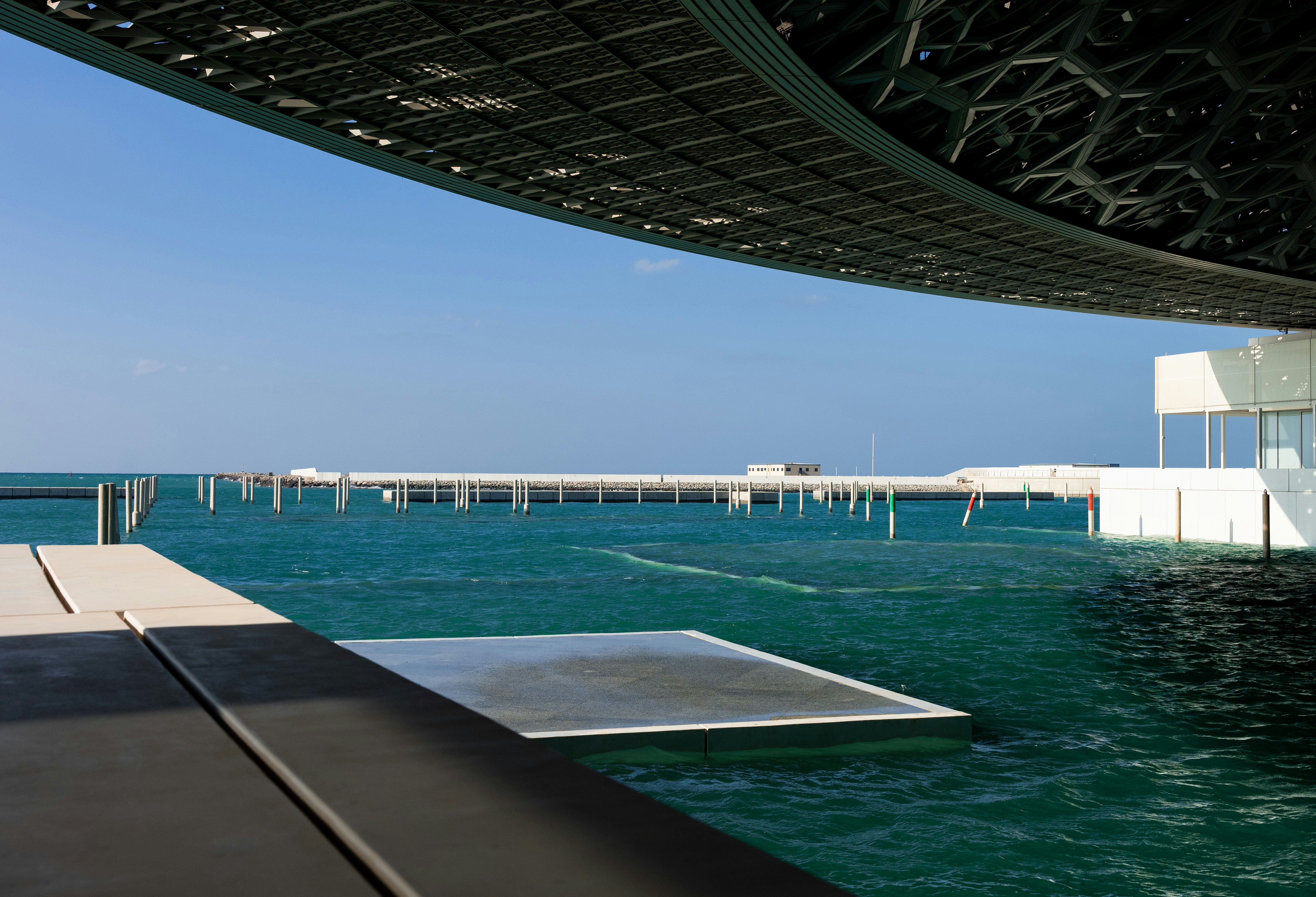 Sunlit waterfront scene featuring a sweeping curved canopy above turquoise water and a series of white platforms extending toward the horizon. Clean architectural lines and a distant pier create a calm, geometric composition.