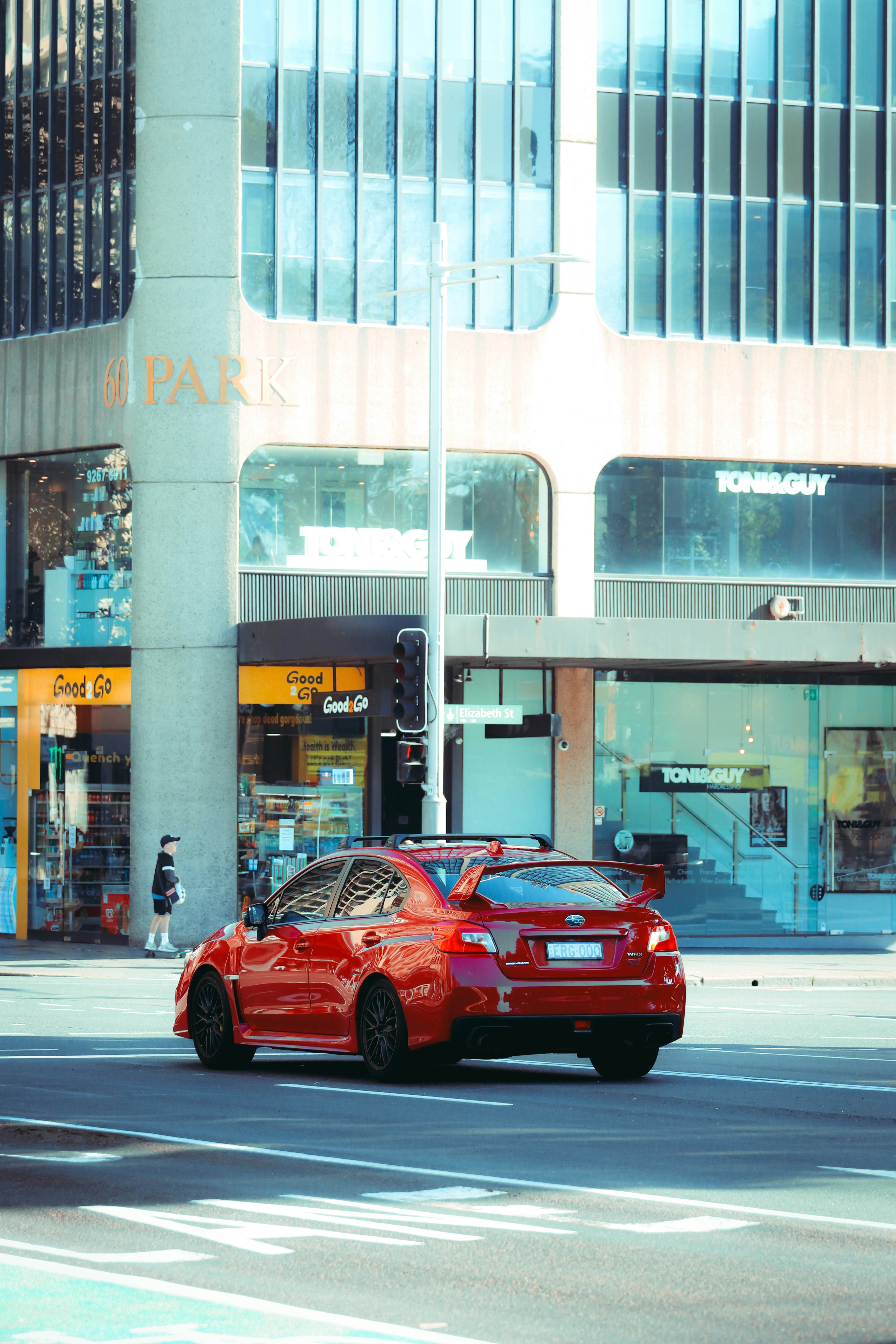 Red sedan anchors a sunlit city block beneath glass-fronted towers