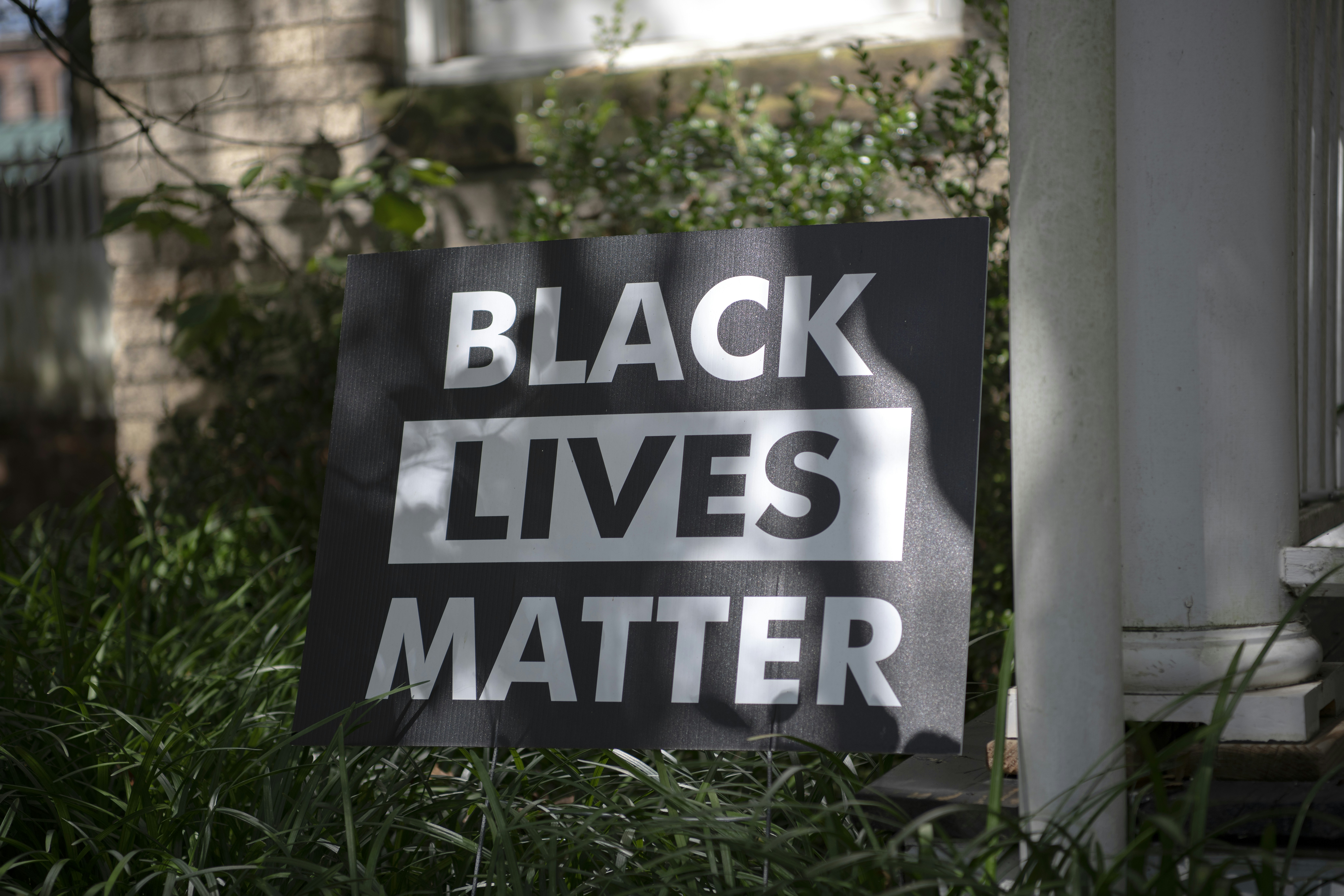 Bold sign reading 'BLACK LIVES MATTER' placed in lush green grass, symbolizing support for social justice. The sunlight creates a play of shadows on the sign.