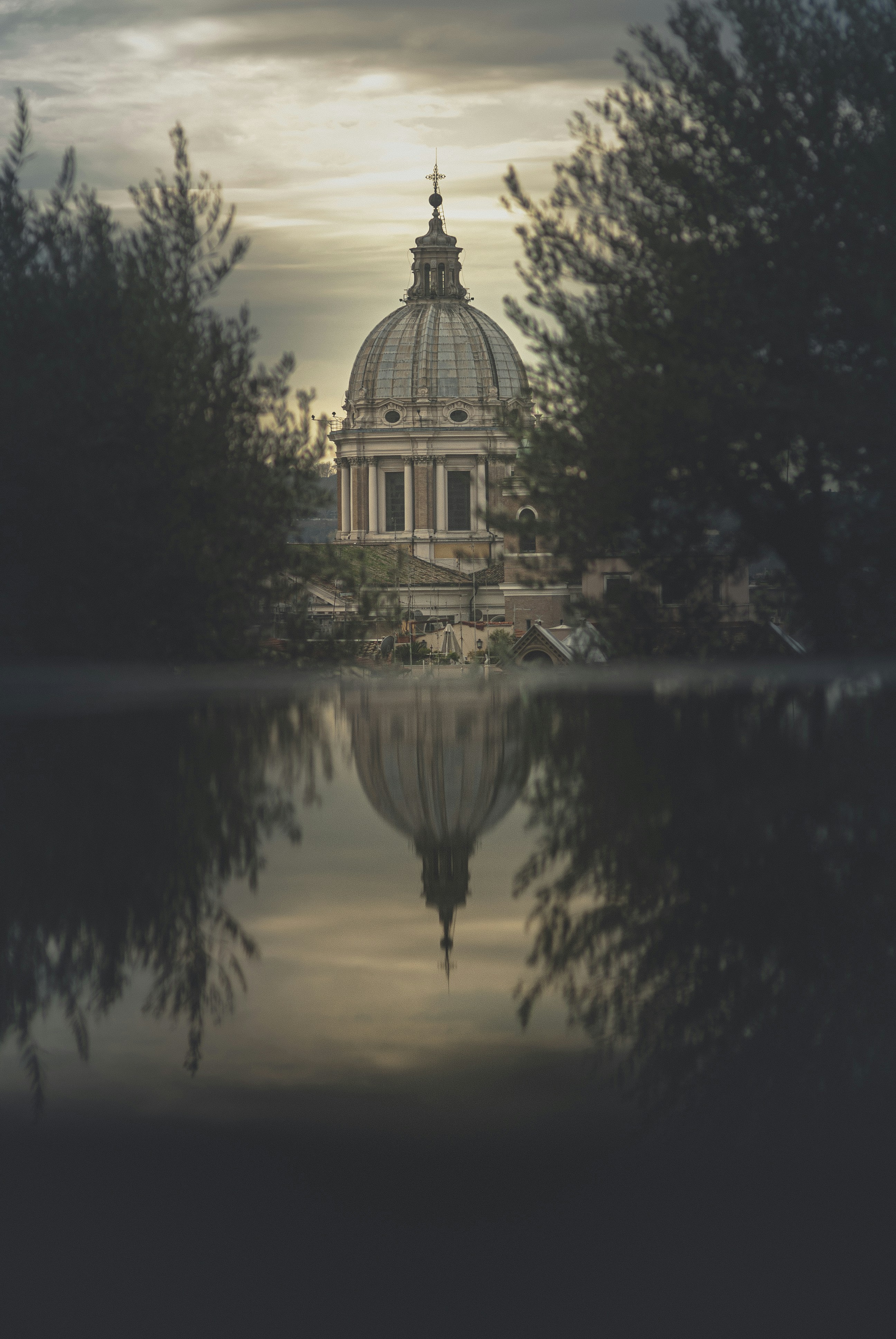 The majestic dome of a historic building is mirrored in still water, framed by surrounding foliage. The scene captures a tranquil moment at dusk.