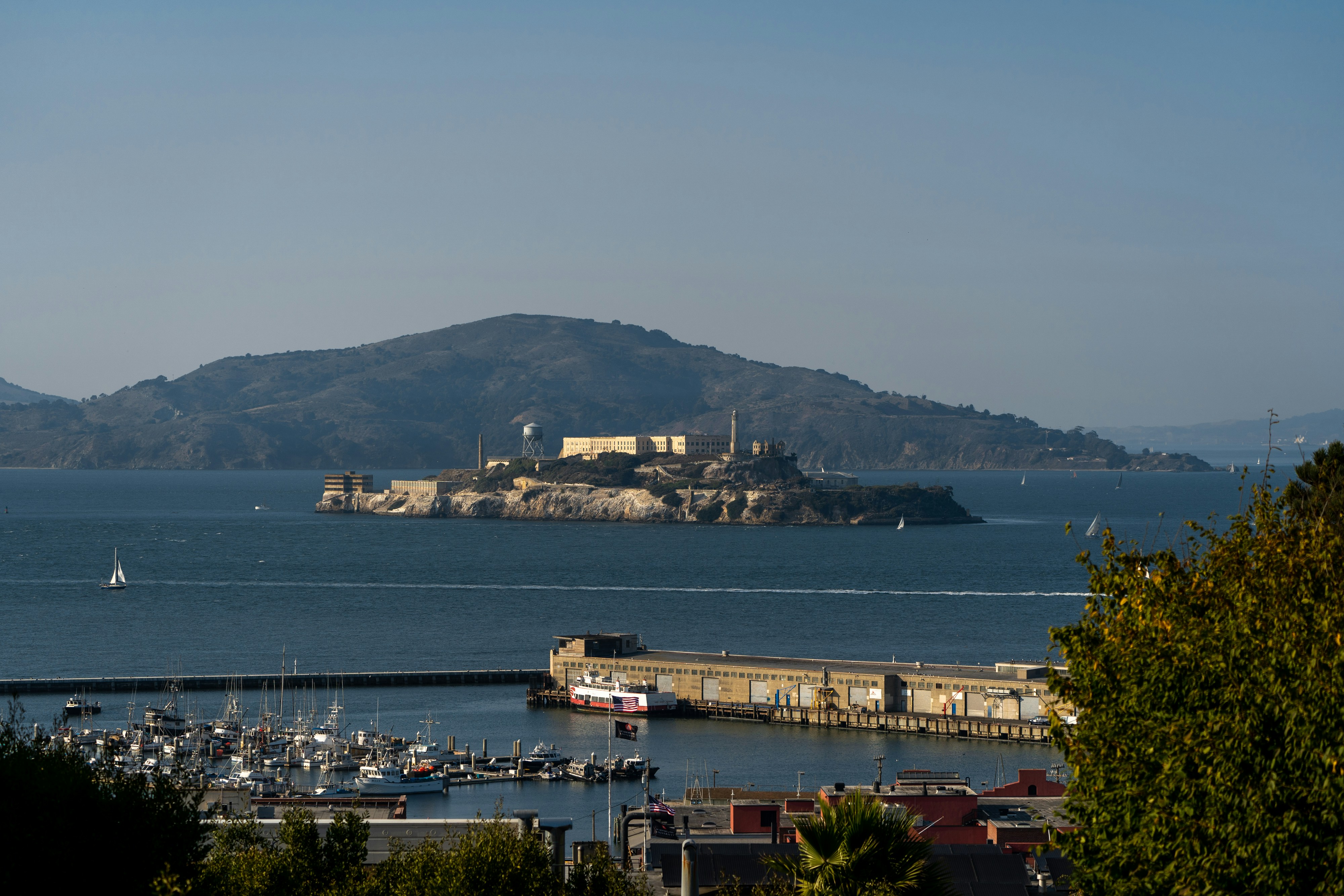 Alcatraz Island rises prominently from the waters, surrounded by a serene bay and distant hills. A marina with boats is visible in the foreground.