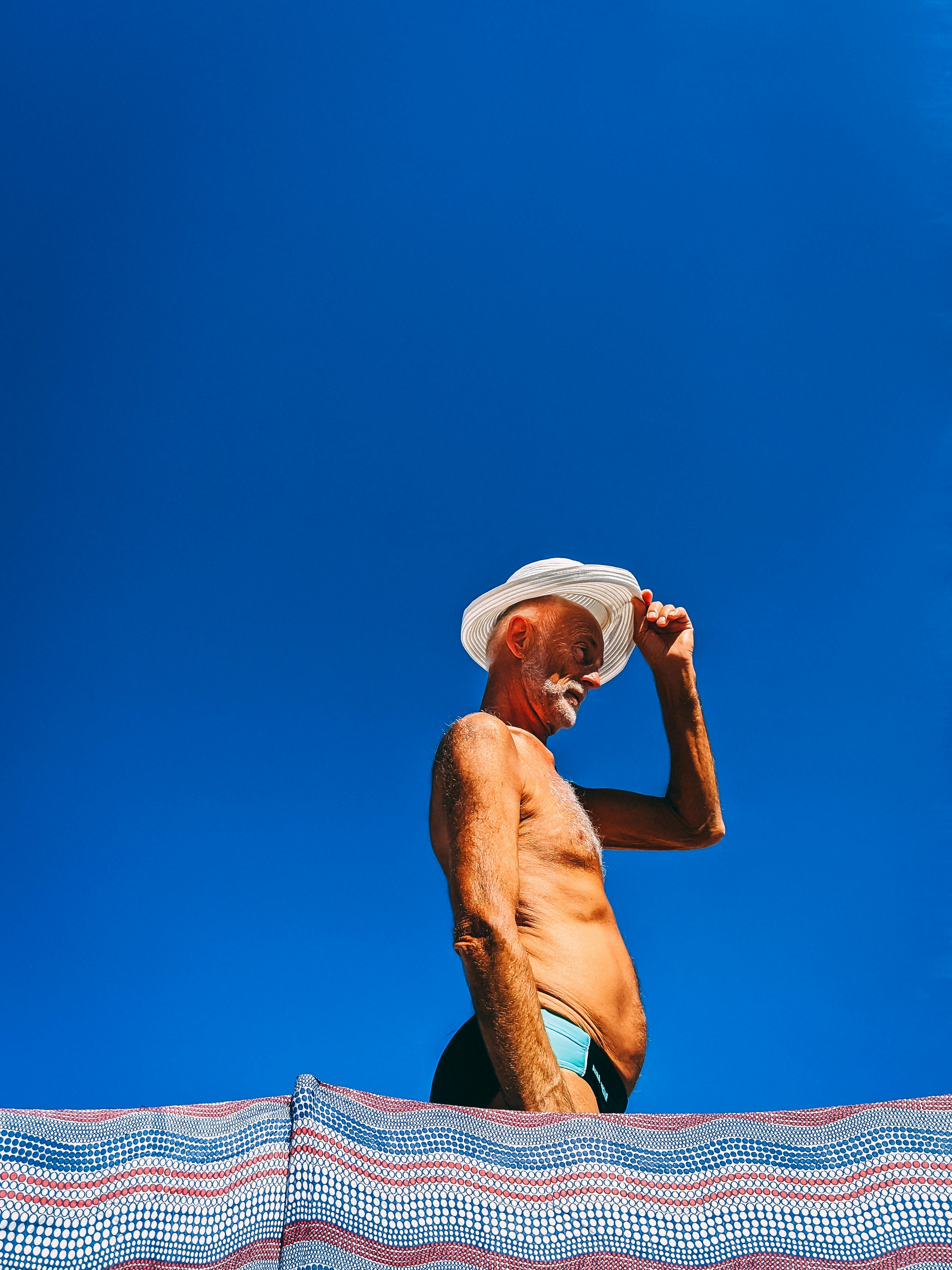 Shirtless older man in a white hat adjusts his brim behind a striped beach towel against a vivid blue sky.