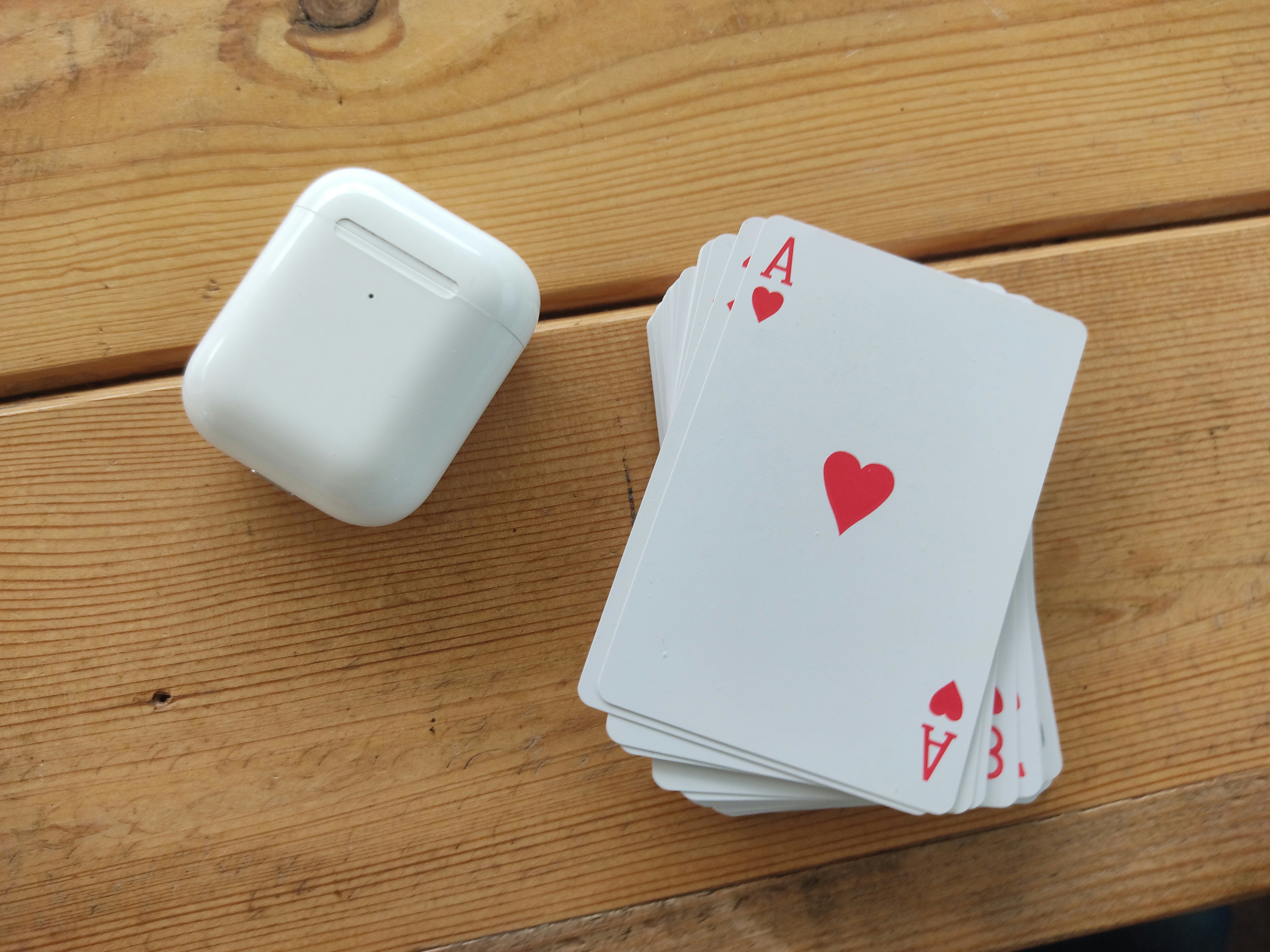 Ace of Hearts from a fanned deck rests on a warm wooden table beside a white wireless earbud case.