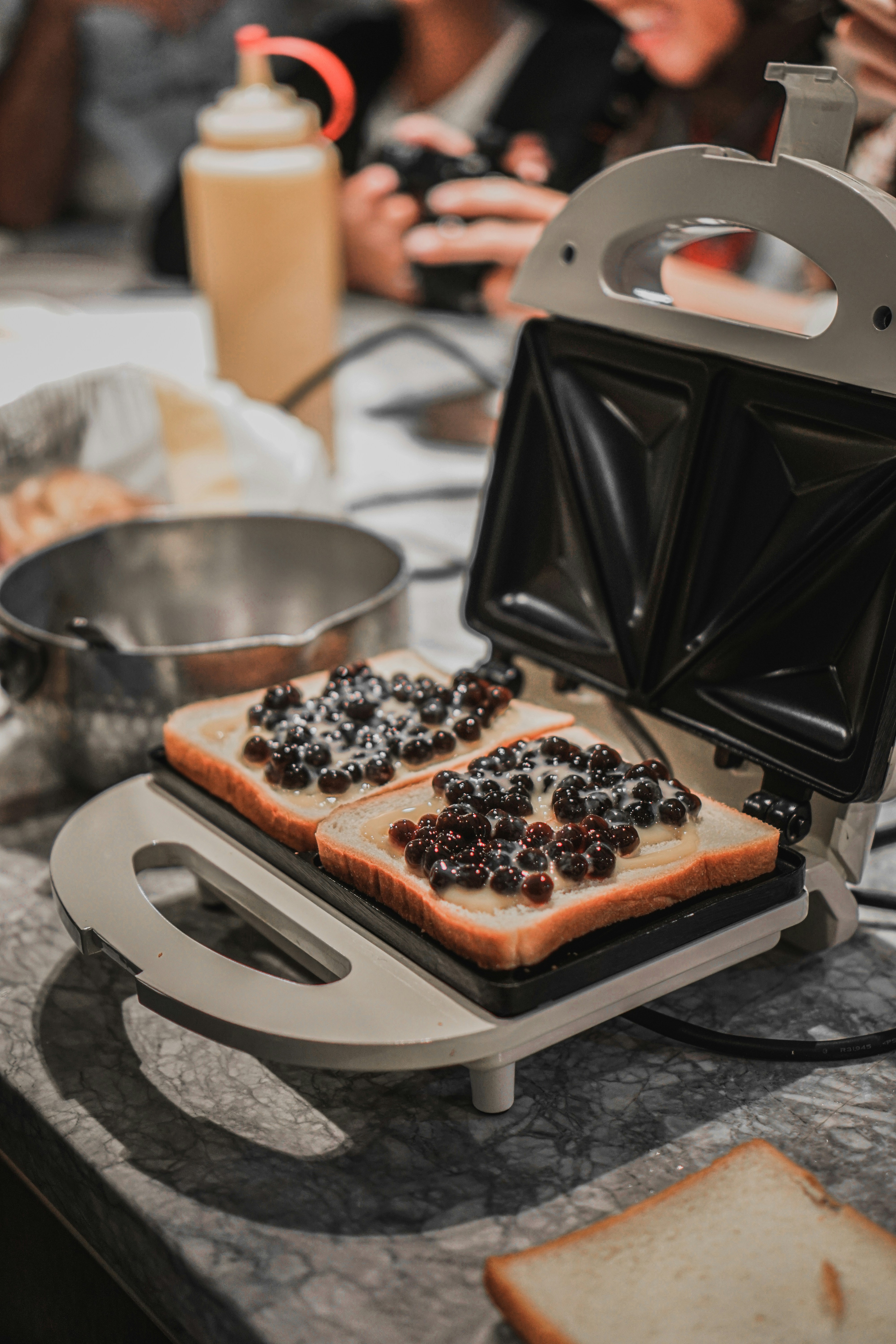A sandwich press holds two slices of bread topped with dark berries, surrounded by kitchen tools and people engaged in cooking.
