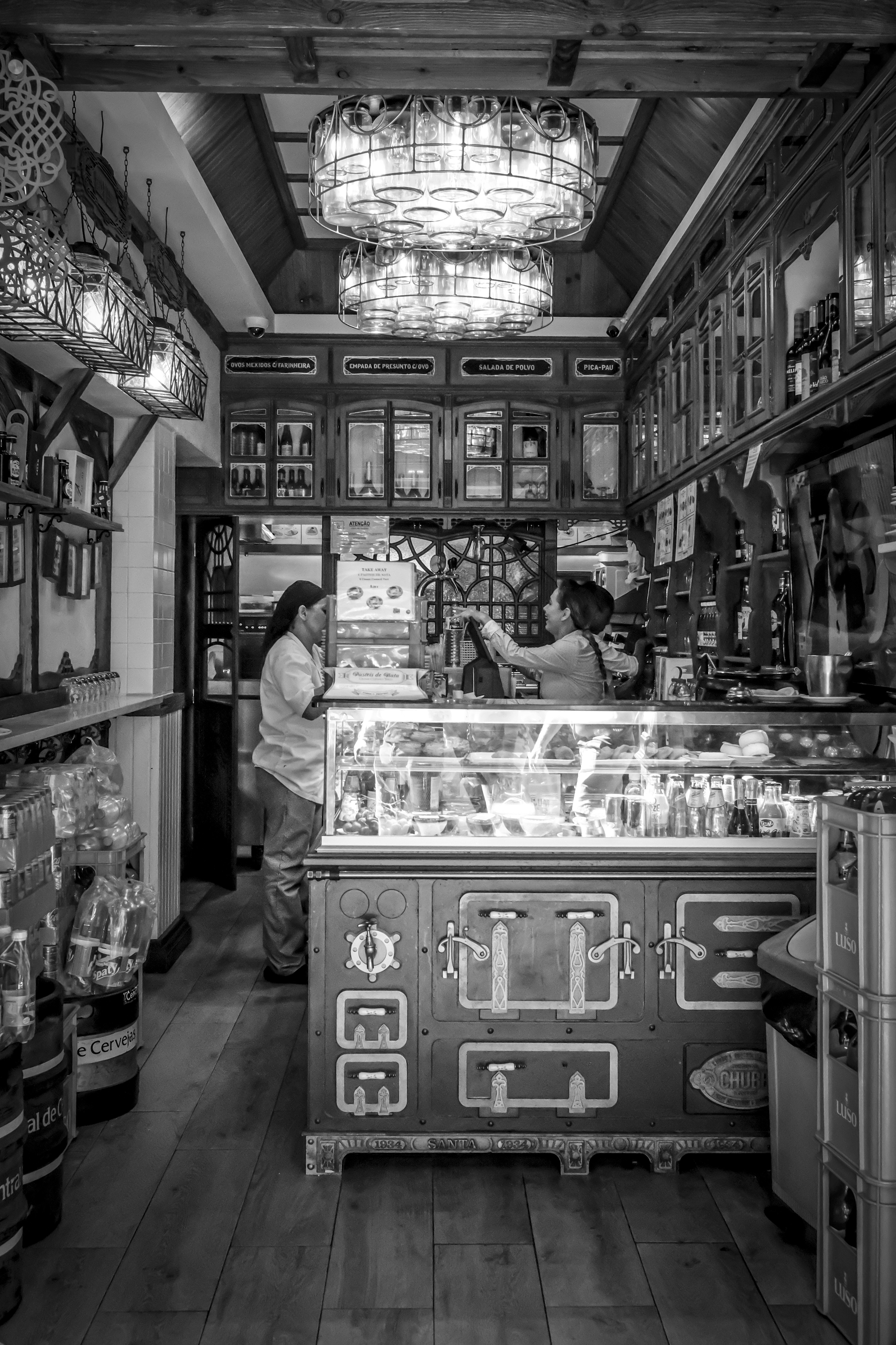 Two baristas engaged in conversation behind a vintage counter filled with drinks, surrounded by rustic decor and warm lighting.