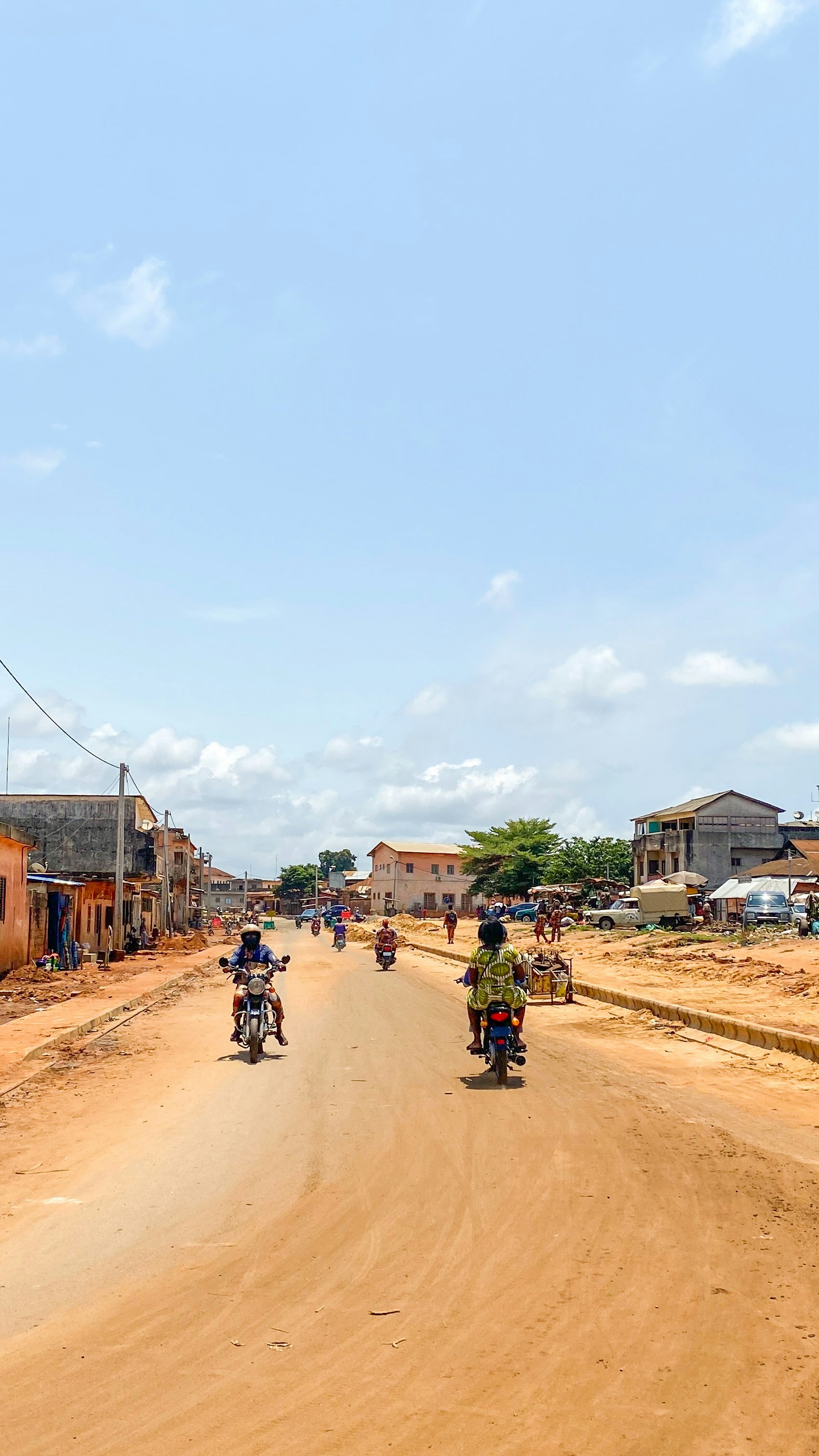 Motorcyclists navigate a sunlit, unpaved road lined with simple buildings and scattered greenery.