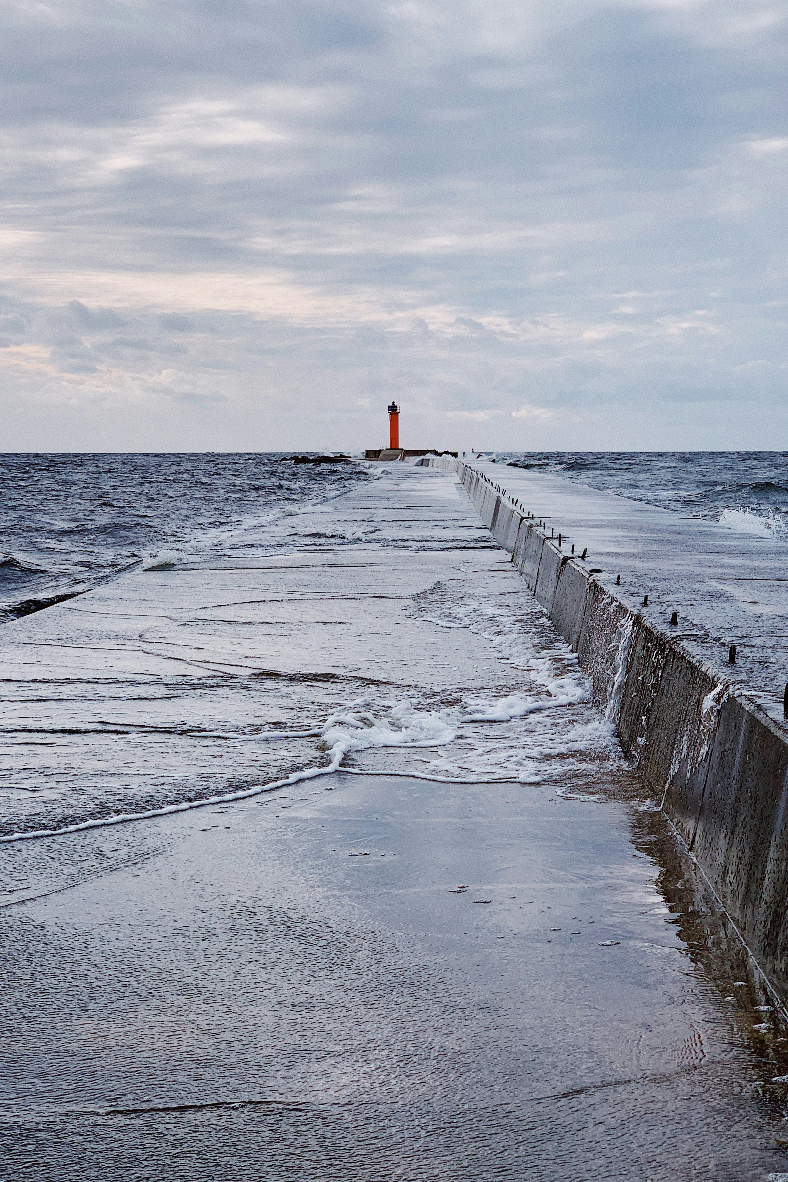 A vibrant orange lighthouse stands tall at the end of a concrete jetty, surrounded by rolling waves under a cloudy sky.