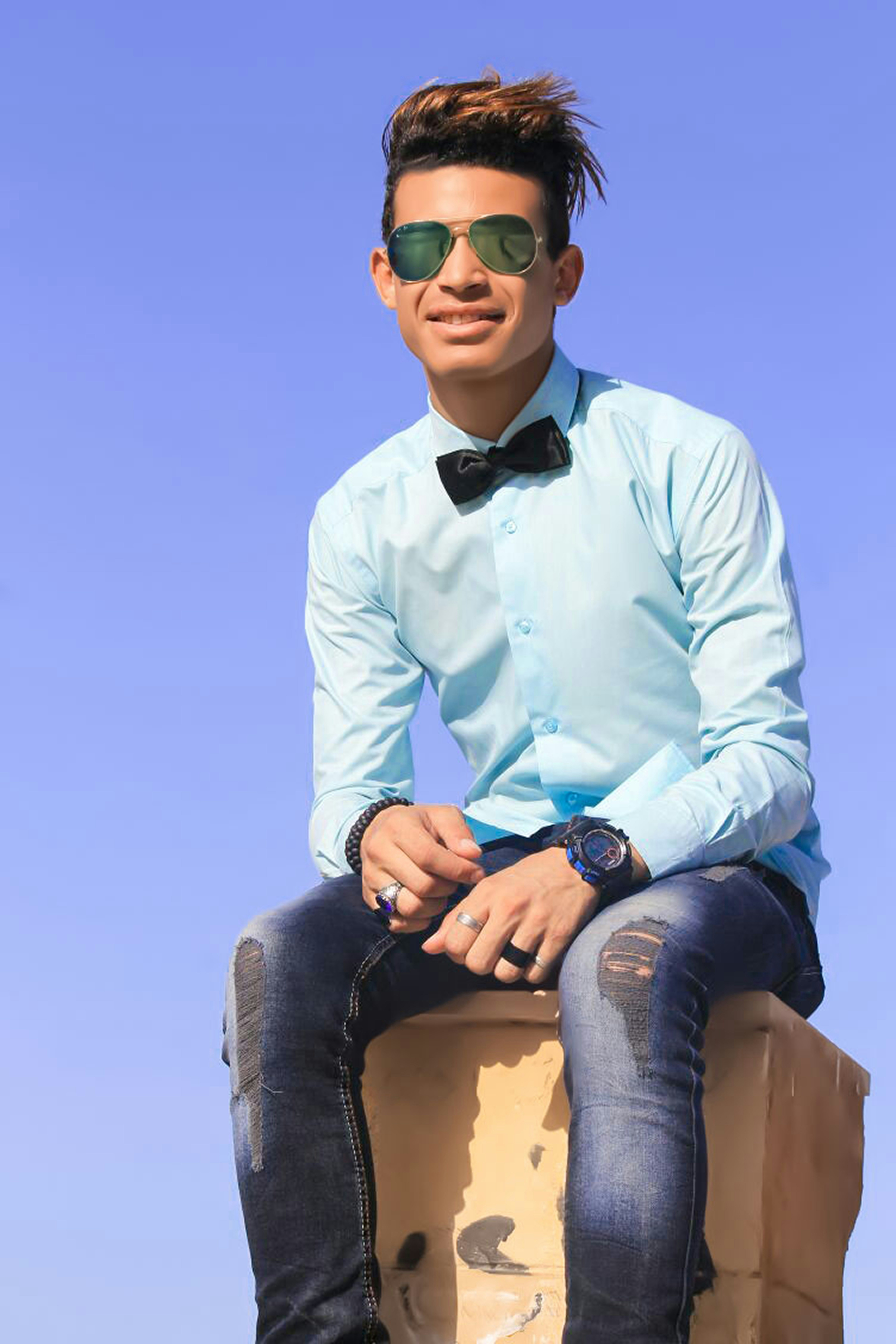 Person in sunglasses and bow tie sitting outdoors with clear blue sky.