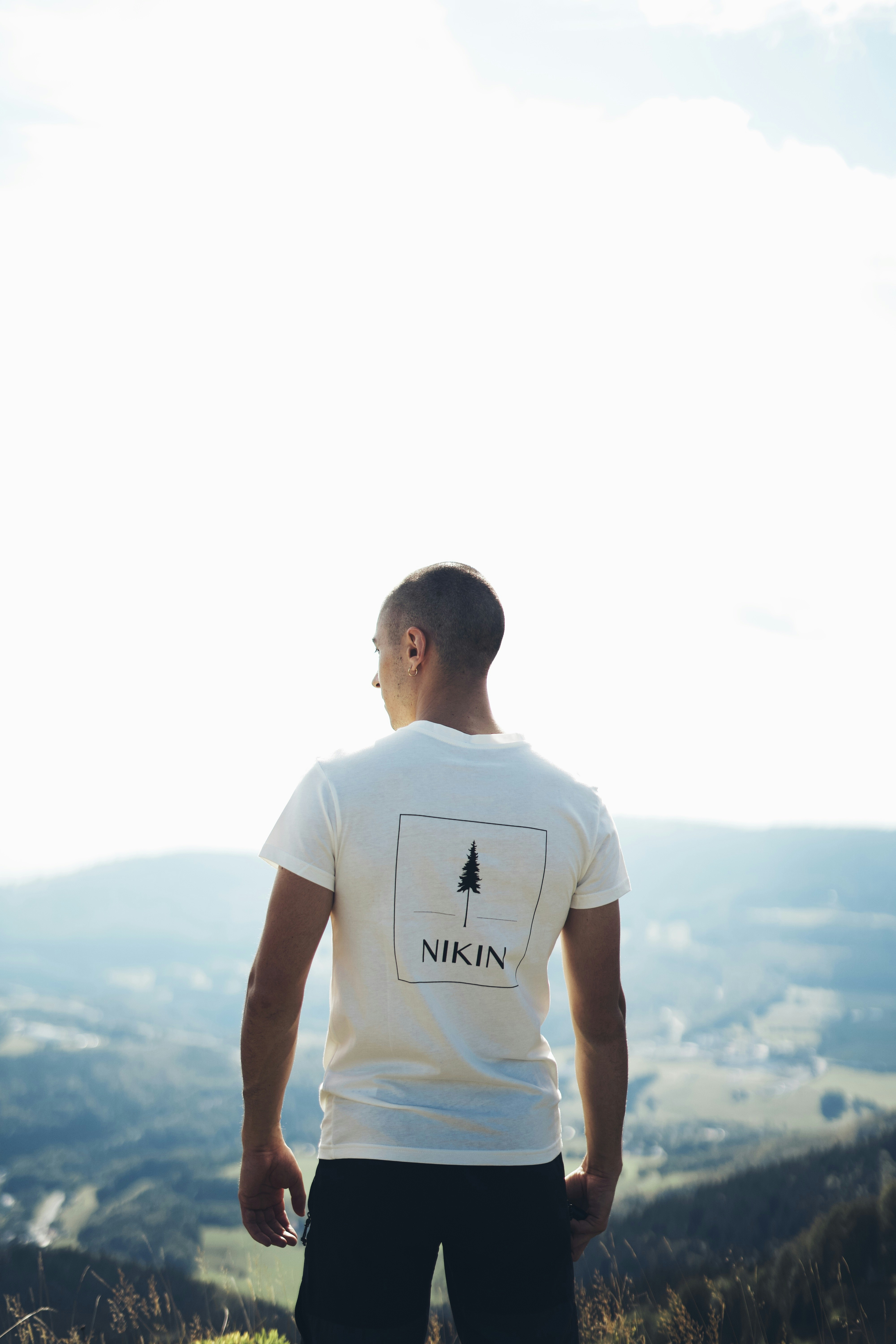 A man stands on a hilltop, gazing into the expansive landscape, wearing a shirt featuring a tree logo. The serene backdrop highlights the connection between humanity and nature.
