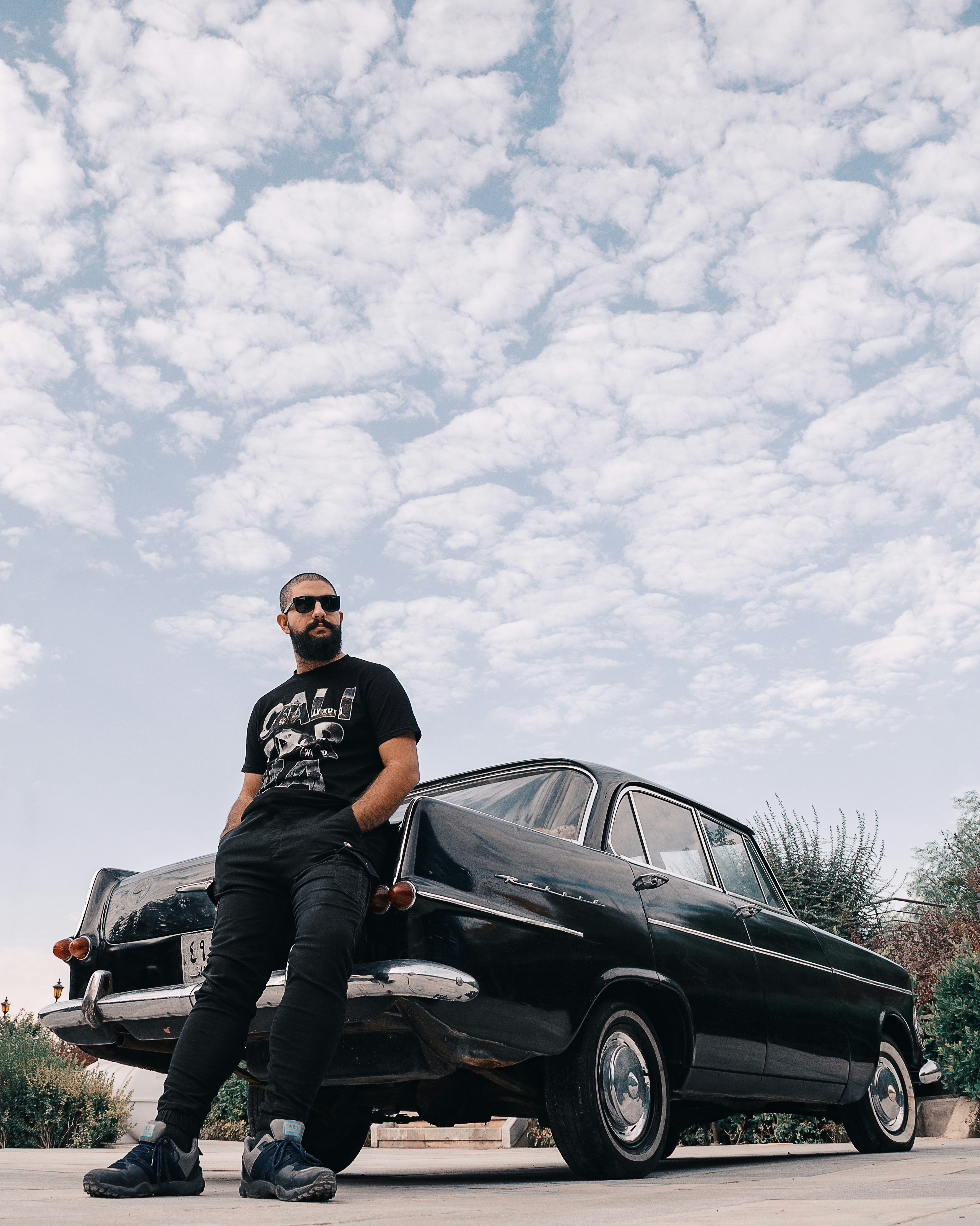 Man in Graphic Tee Leans Against a Retro Car Under a Sky of Cotton Clouds