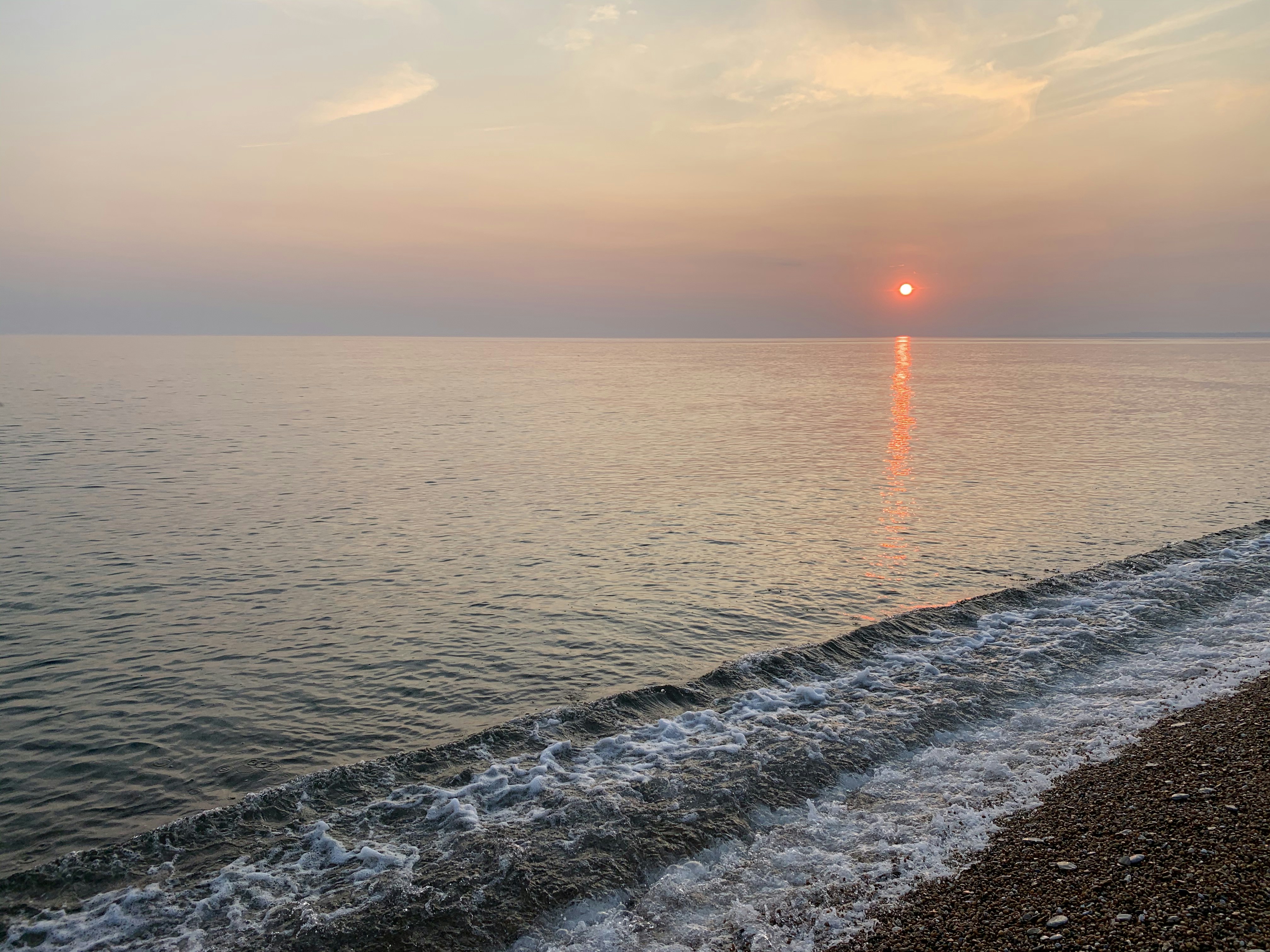 Gentle waves lap against a pebble-strewn shore as the sun sets over a calm sea, casting a warm glow.