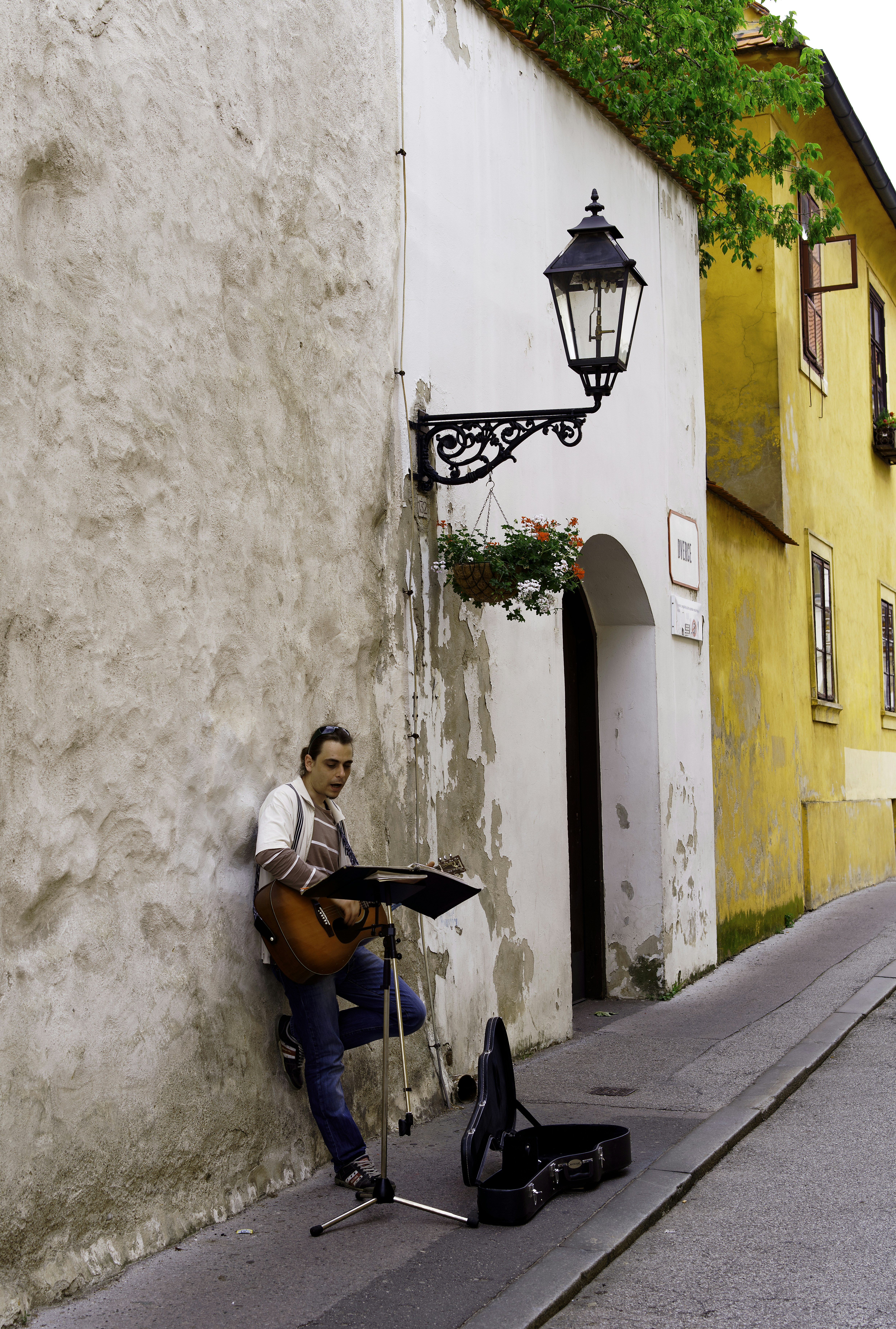 Musician playing guitar against a textured wall in a quaint alley, with a lantern and vibrant buildings adding charm.