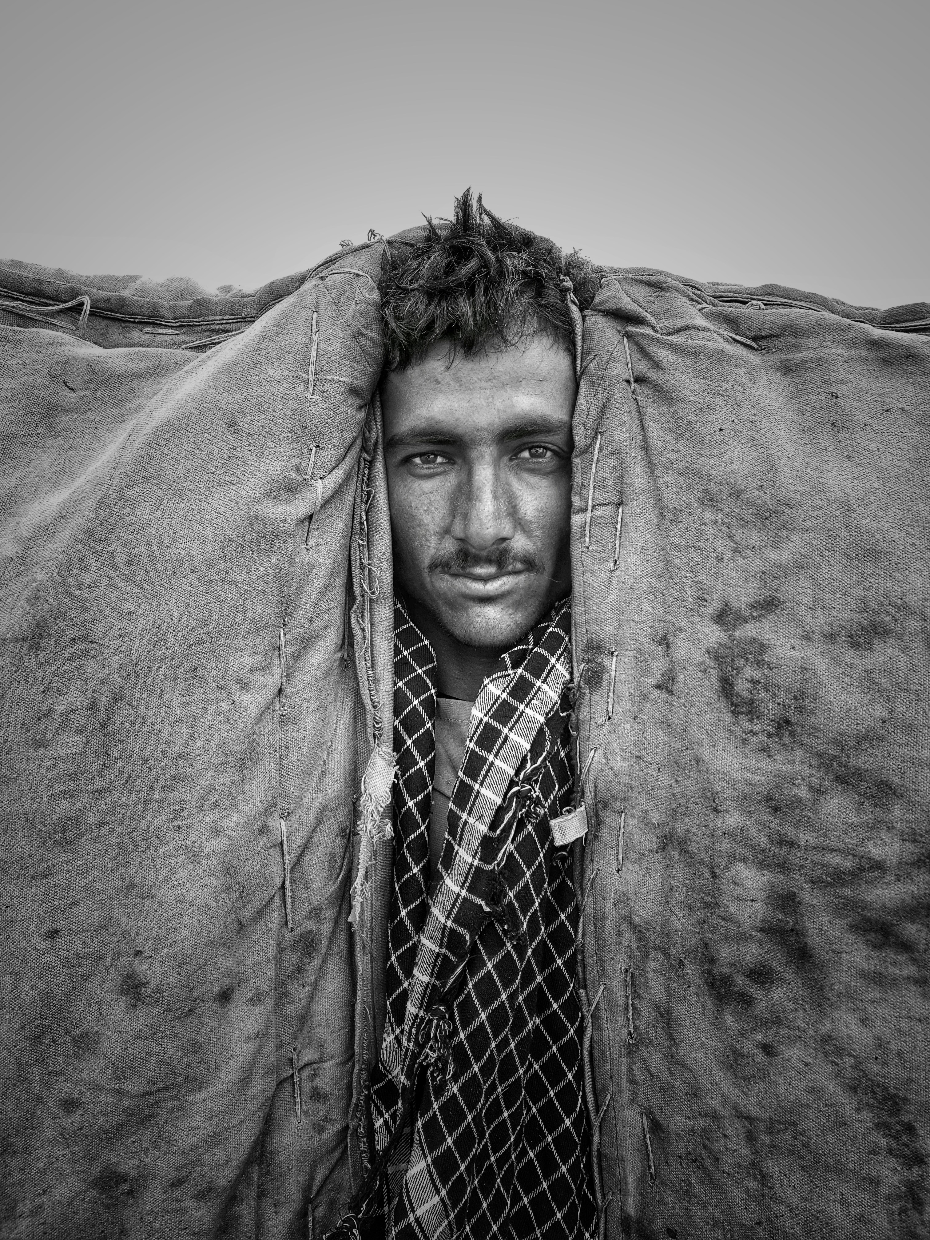 Photograph of a man peering through a rough textile shelter, wrapped in a checkered scarf.