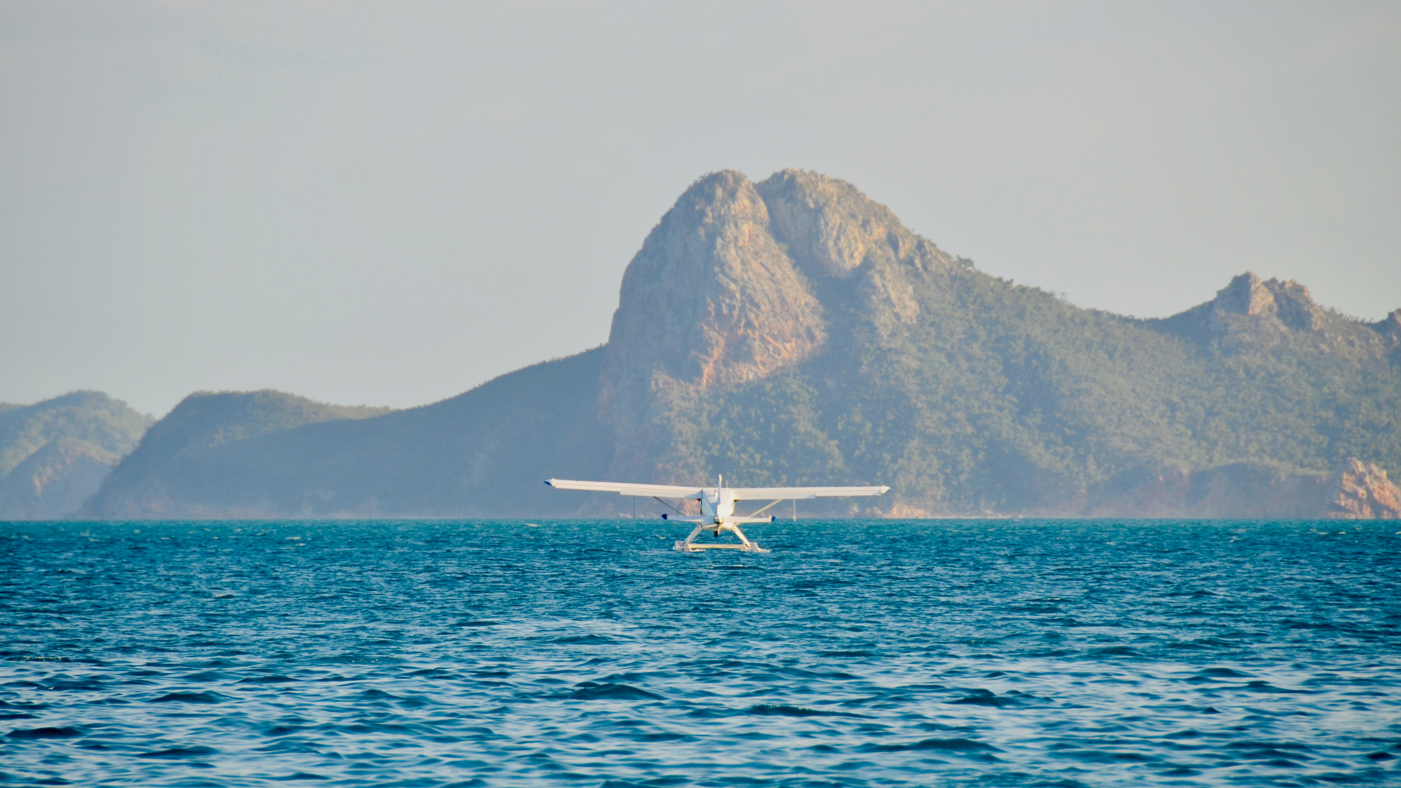 A seaplane glides over calm blue water with rugged mountains in the background, captured in a photograph.