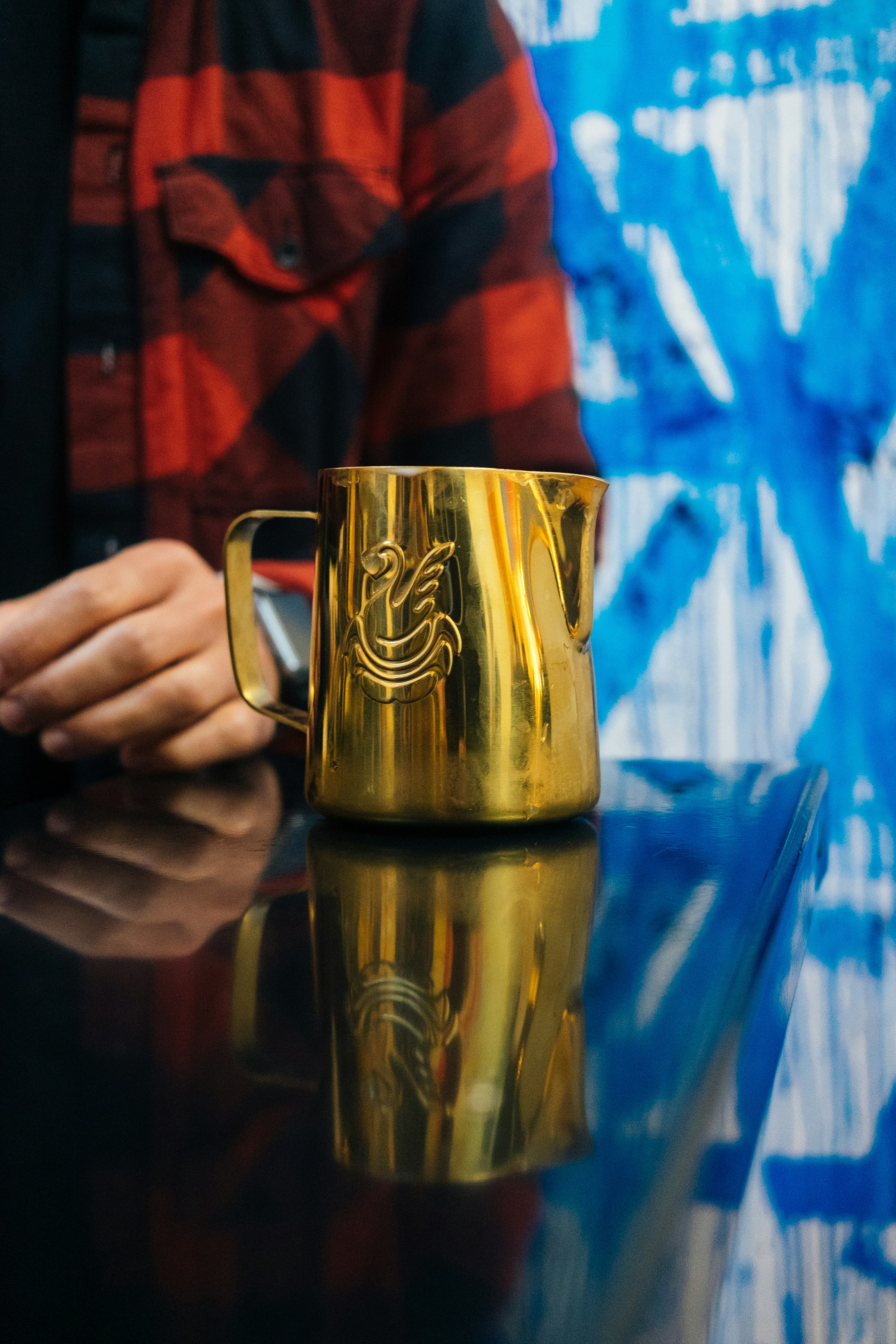 Golden milk frothing pitcher rests on a reflective surface, with a blurred background featuring a vibrant blue mural.